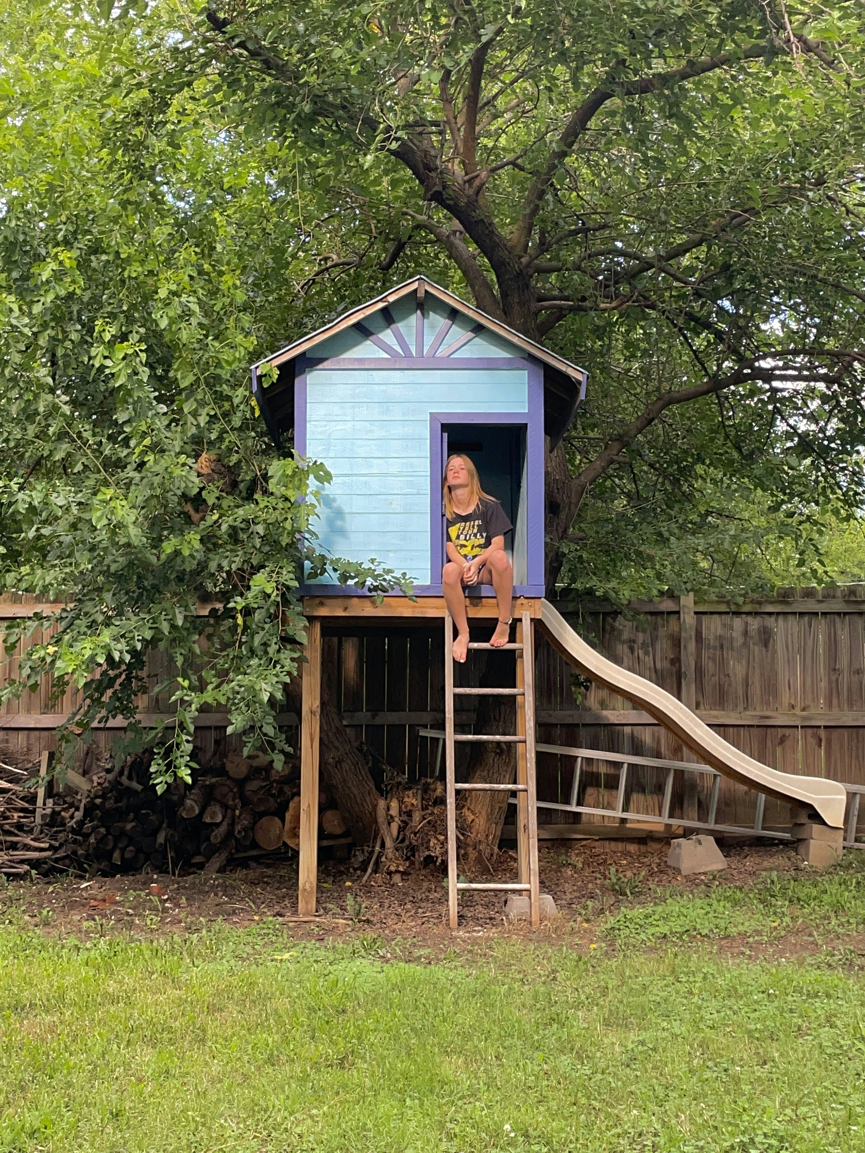 A child relaxes in a colorful treehouse, framed by lush greenery and a wooden fence. The slide adds a playful touch to this backyard escape.