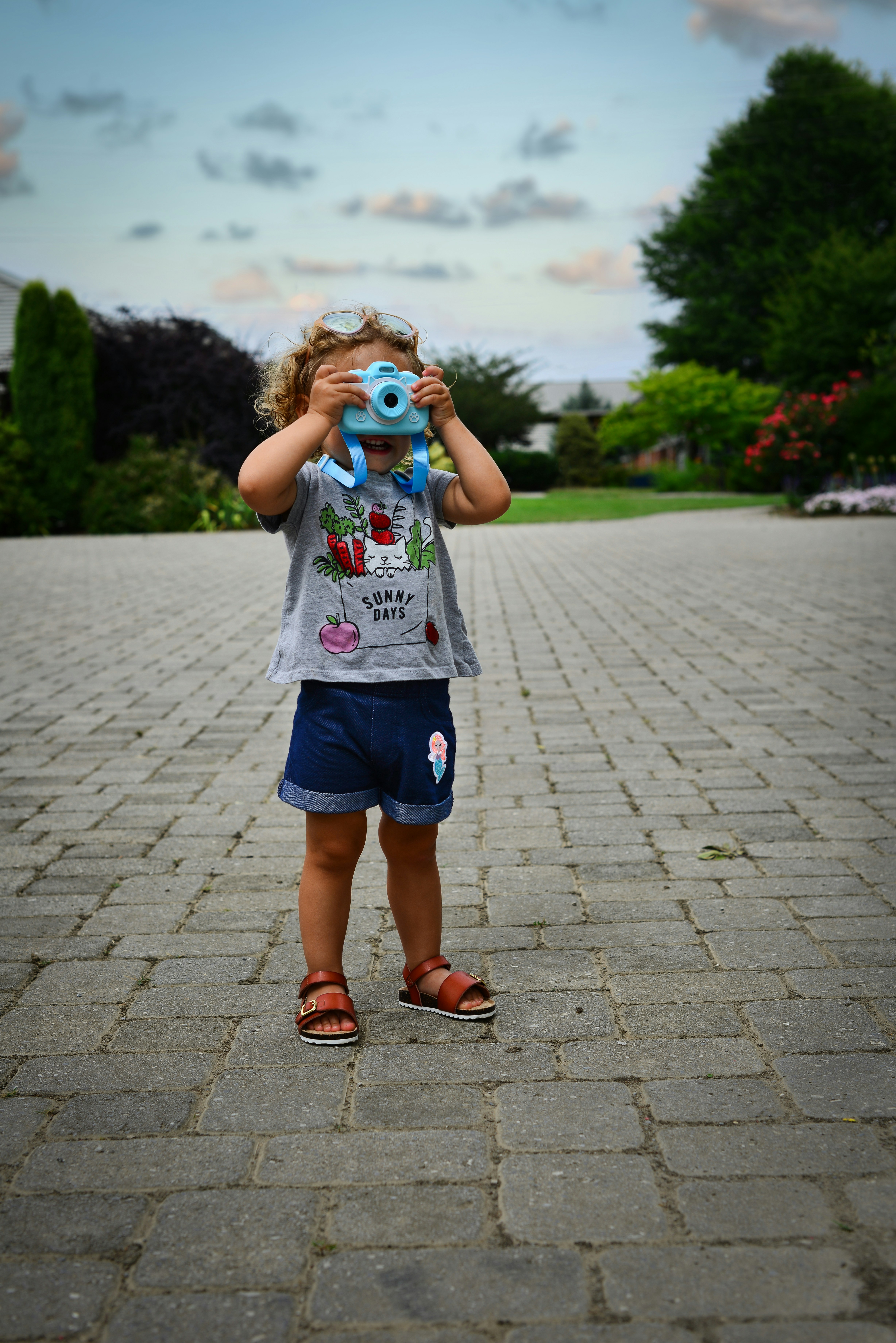 Child holding a toy camera, standing on a stone pathway surrounded by greenery and flowers.