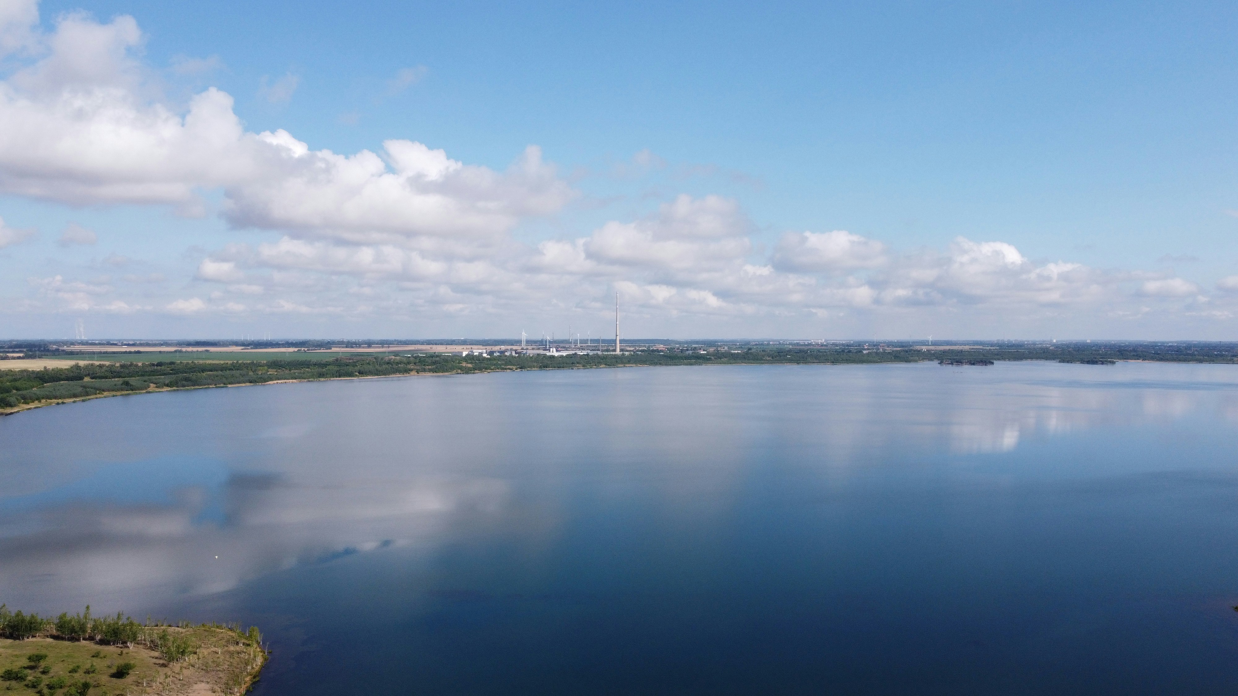 Expansive view of a tranquil lake reflecting clouds and distant industrial structures. The scene captures the juxtaposition of nature and human activity.