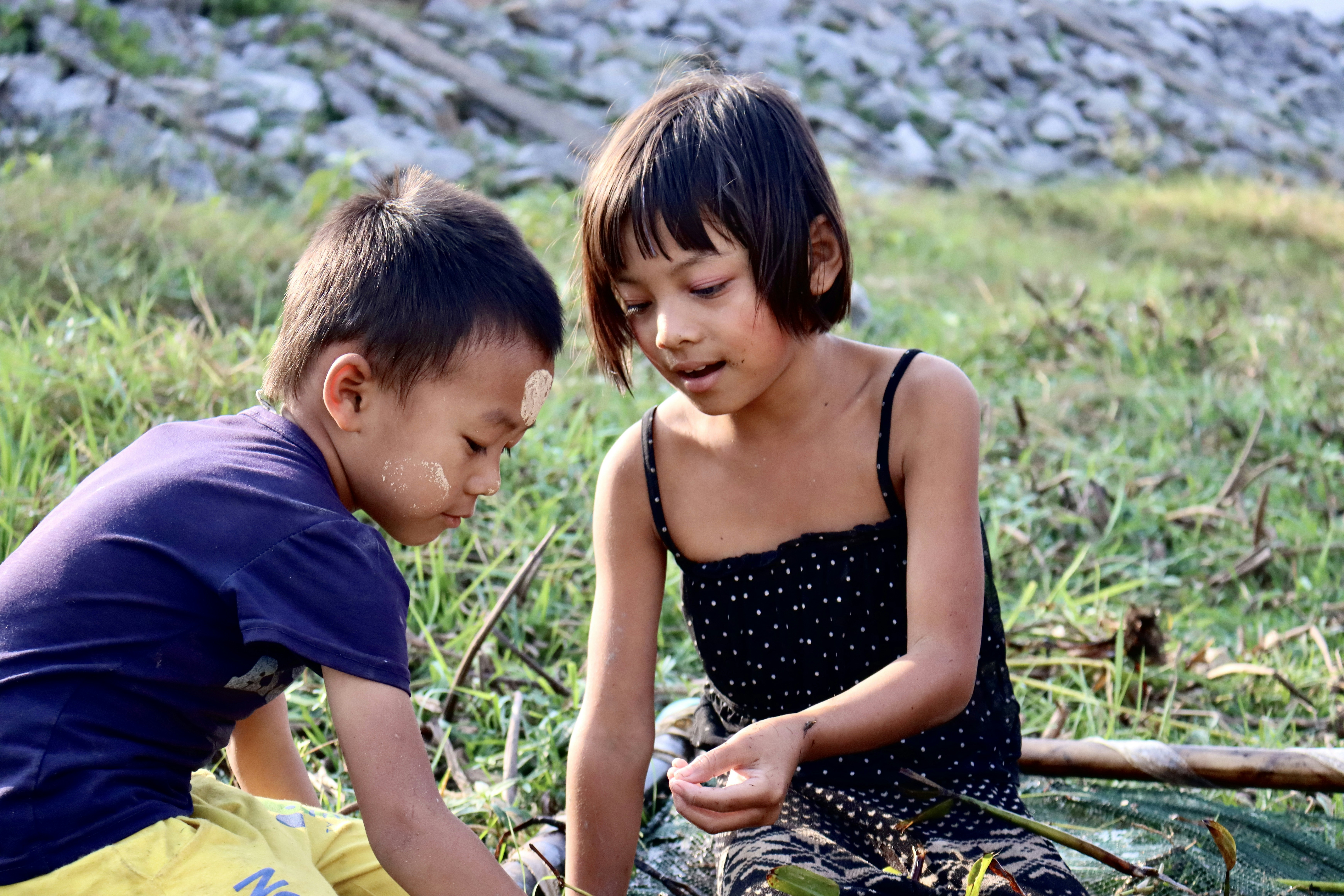 Two children engaged in playful exploration on a grassy patch, surrounded by natural elements. The scene captures their curiosity and connection to the outdoors.
