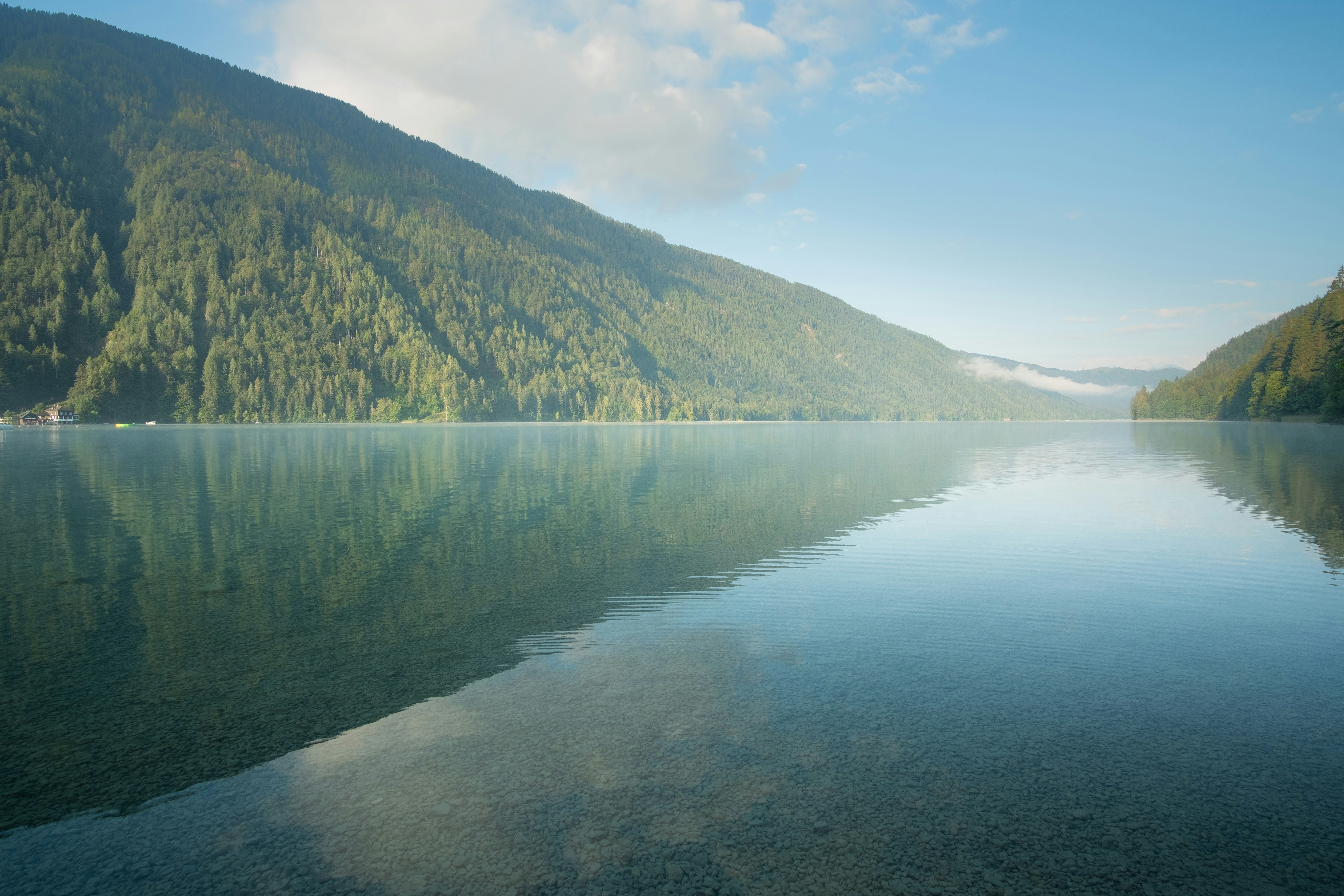Clear turquoise waters reflect lush green hills under a bright morning sky at Lake Weissensee.
