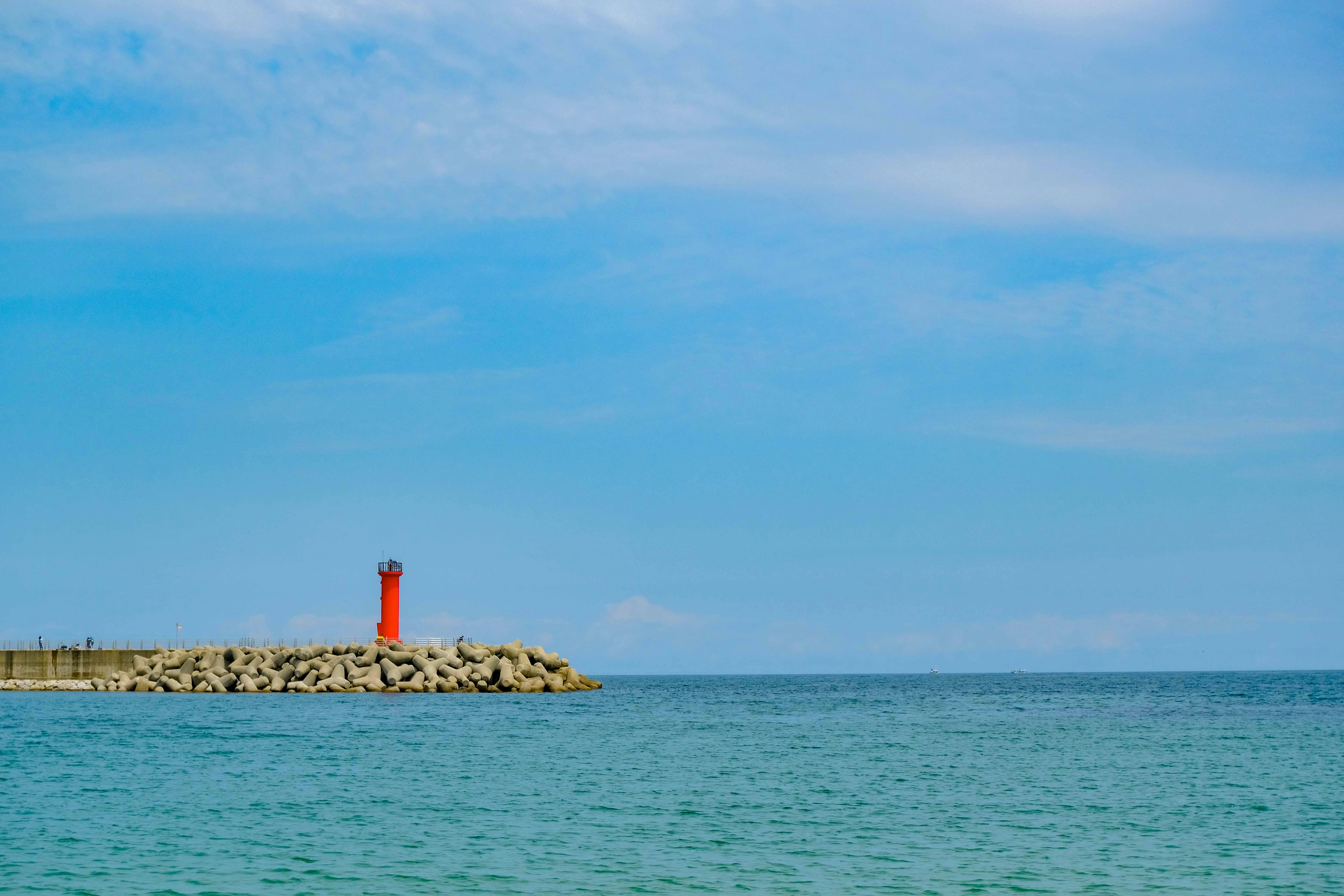 Bright red lighthouse standing tall at the edge of a rocky pier, overlooking calm blue waters under a clear sky.