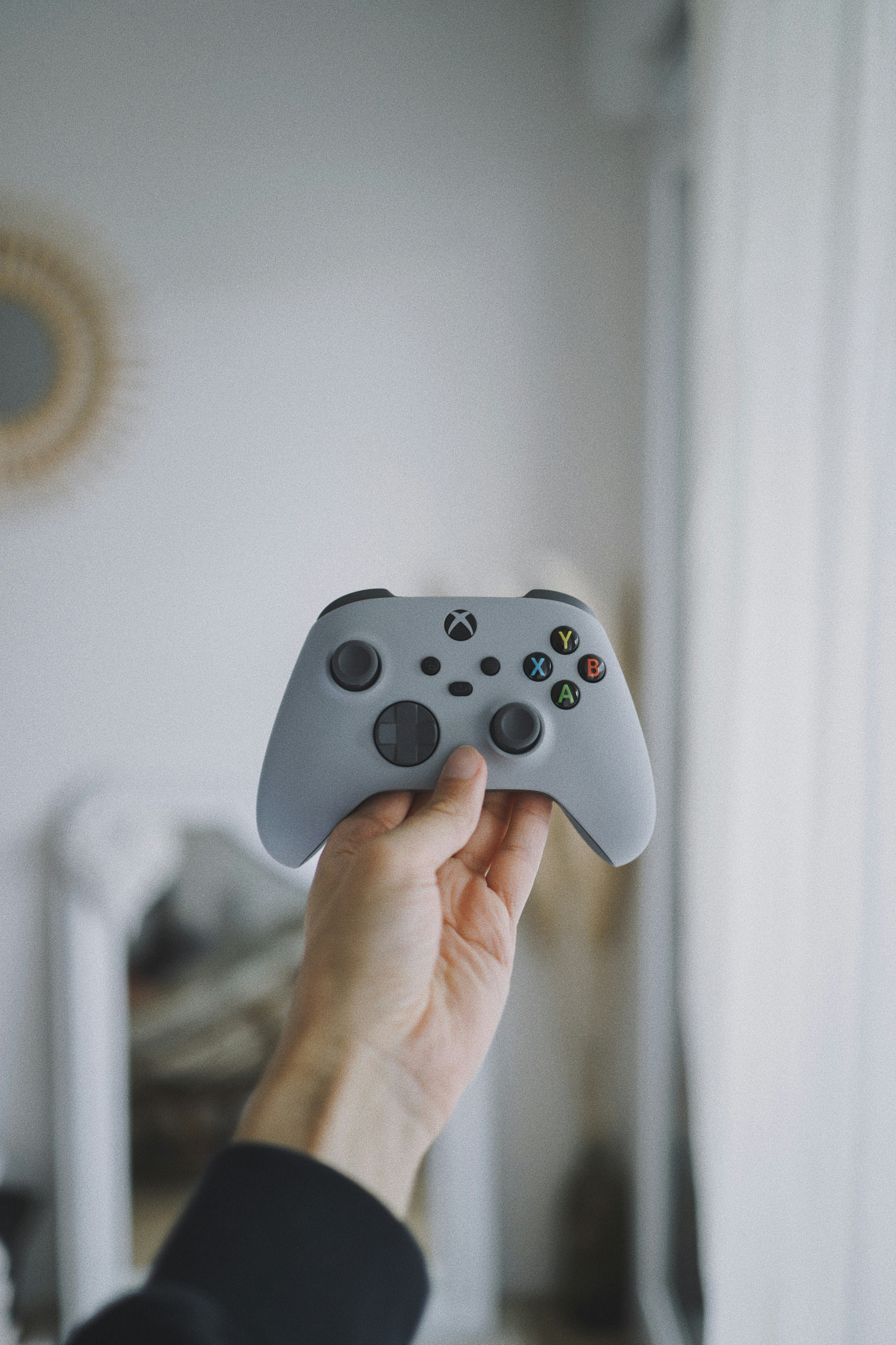 A hand holds a gray Xbox controller against a softly blurred background, showcasing a blend of comfort and technology. The scene reflects a cozy gaming environment.