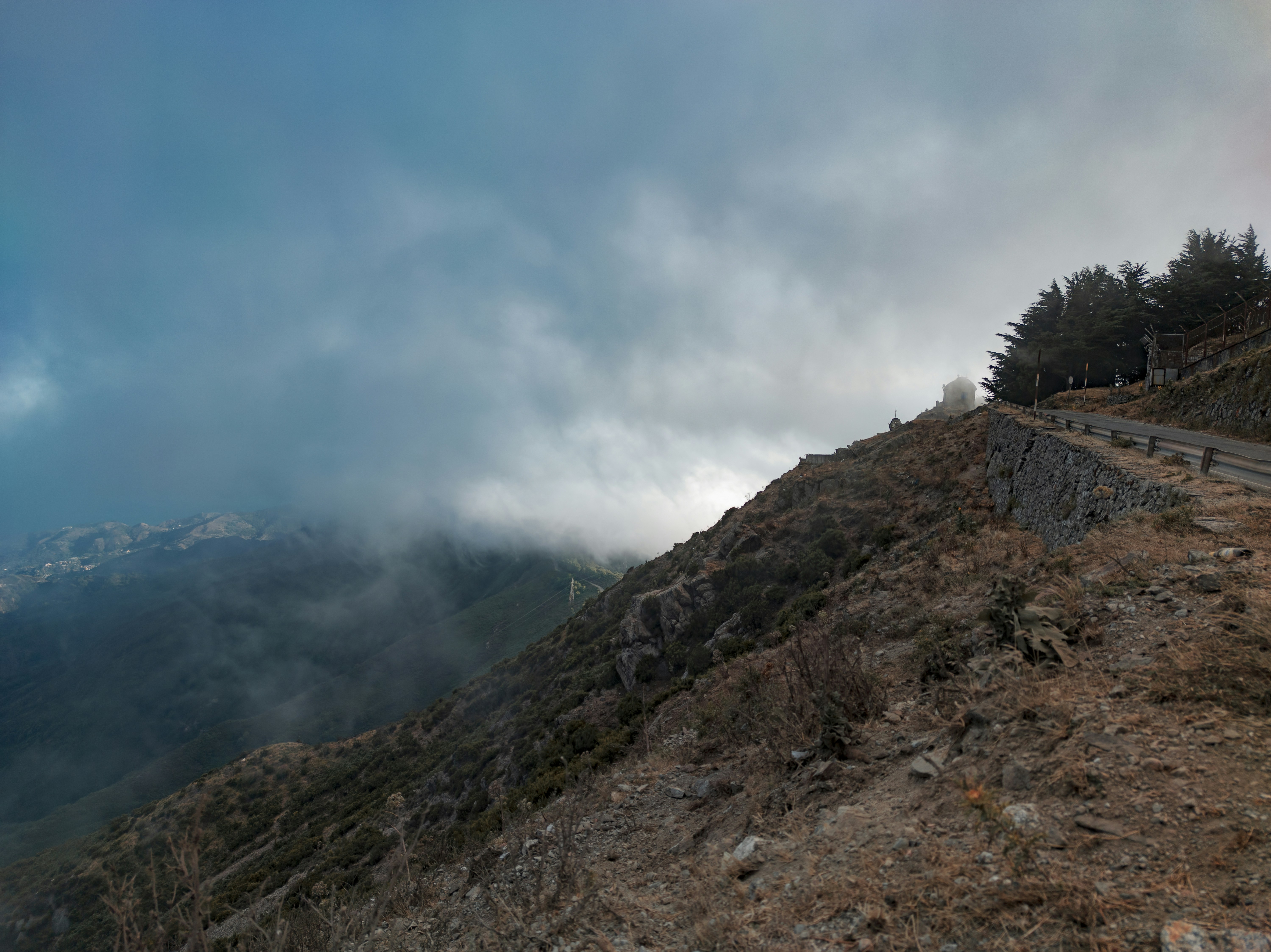 Fog envelops a winding mountain road, revealing a rugged landscape dotted with sparse vegetation. The scene captures the serene yet mysterious atmosphere of high altitudes.