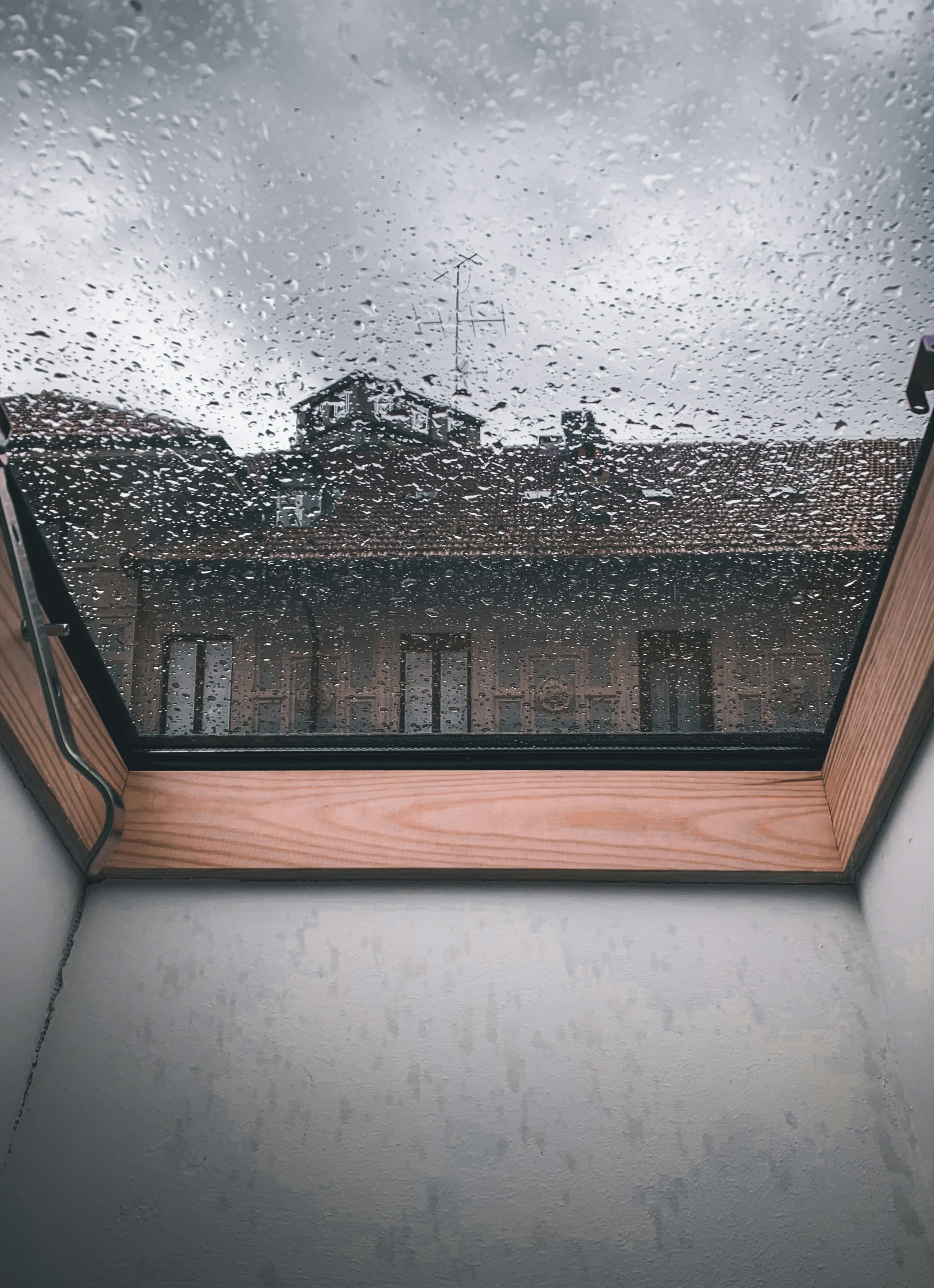Rain-soaked skylight revealing a textured view of rooftops under a cloudy sky. The droplets create a natural filter, enhancing the scene's depth.