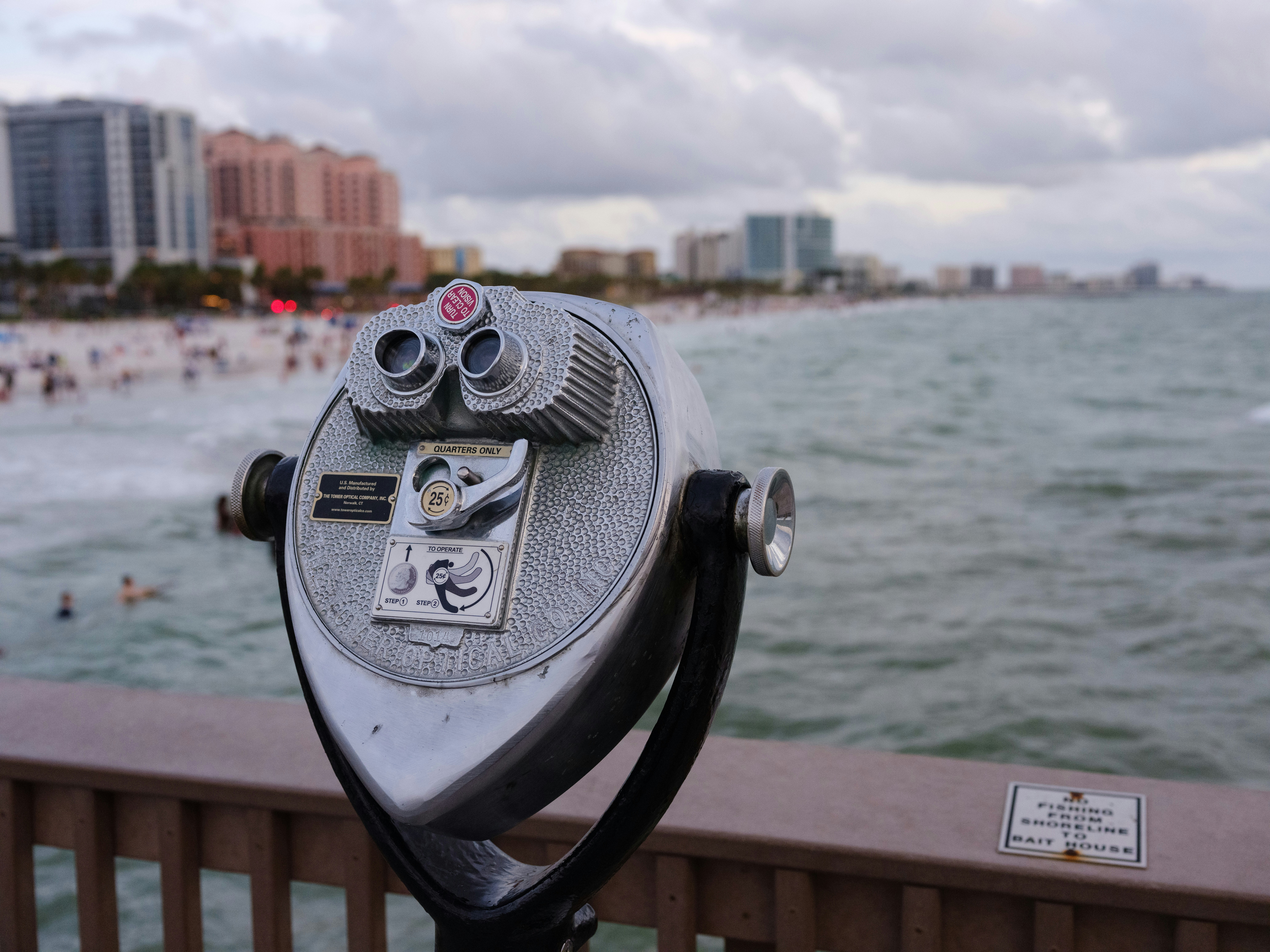 Coin-operated binoculars on a pier overlooking the ocean with a distant cityscape under a cloudy sky.