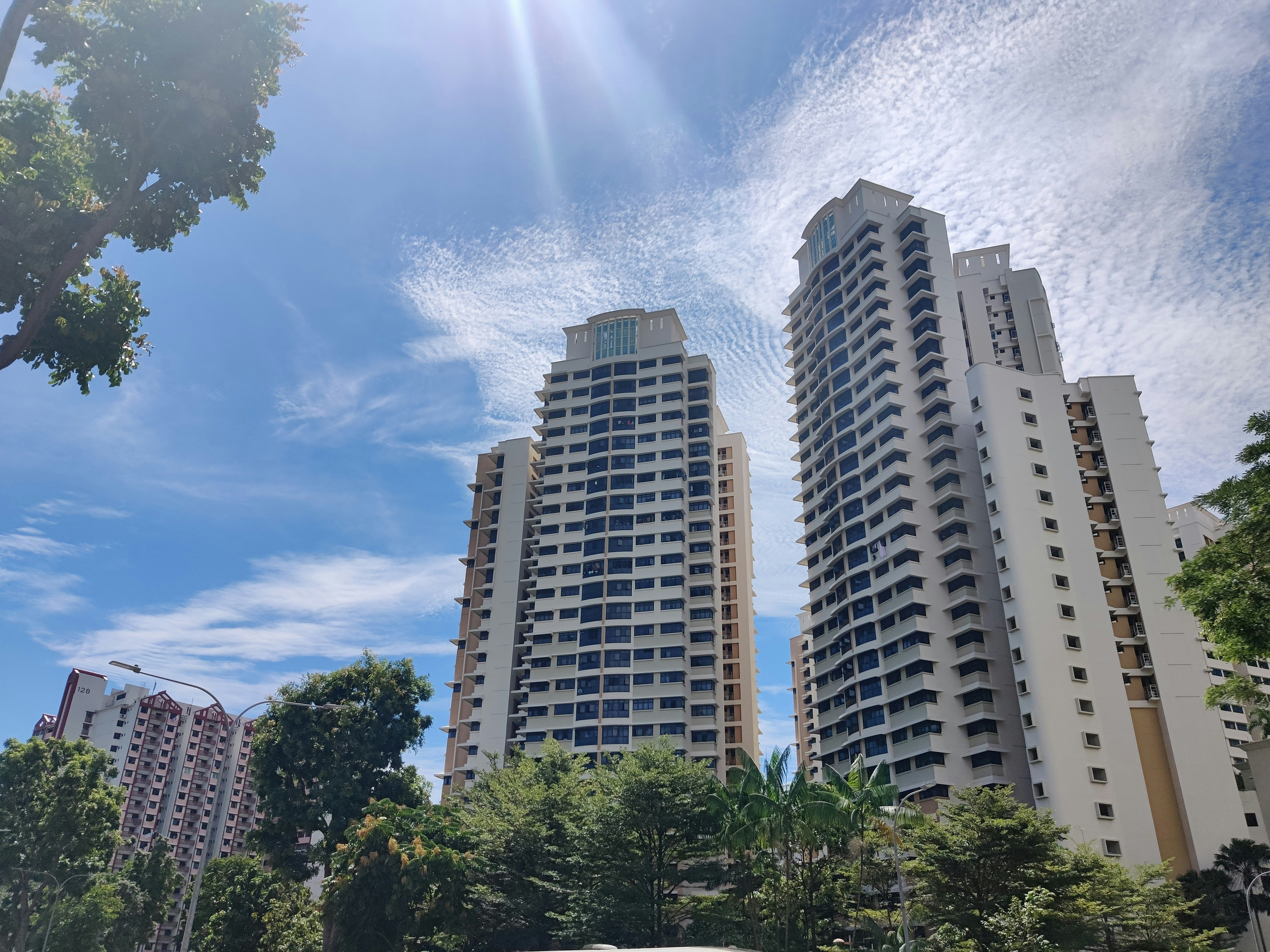 Tall residential buildings rise against a backdrop of wispy clouds and bright blue sky, framed by lush greenery below.