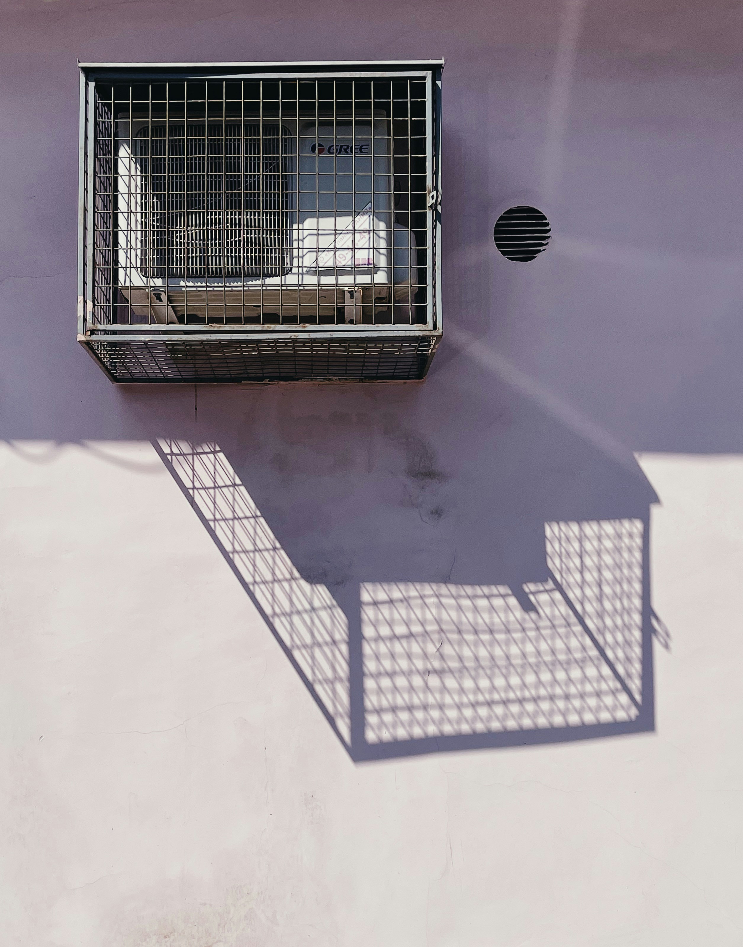 Lilac Wall, Gridded Shade: An Outdoor AC Cage and Its Looming Shadow