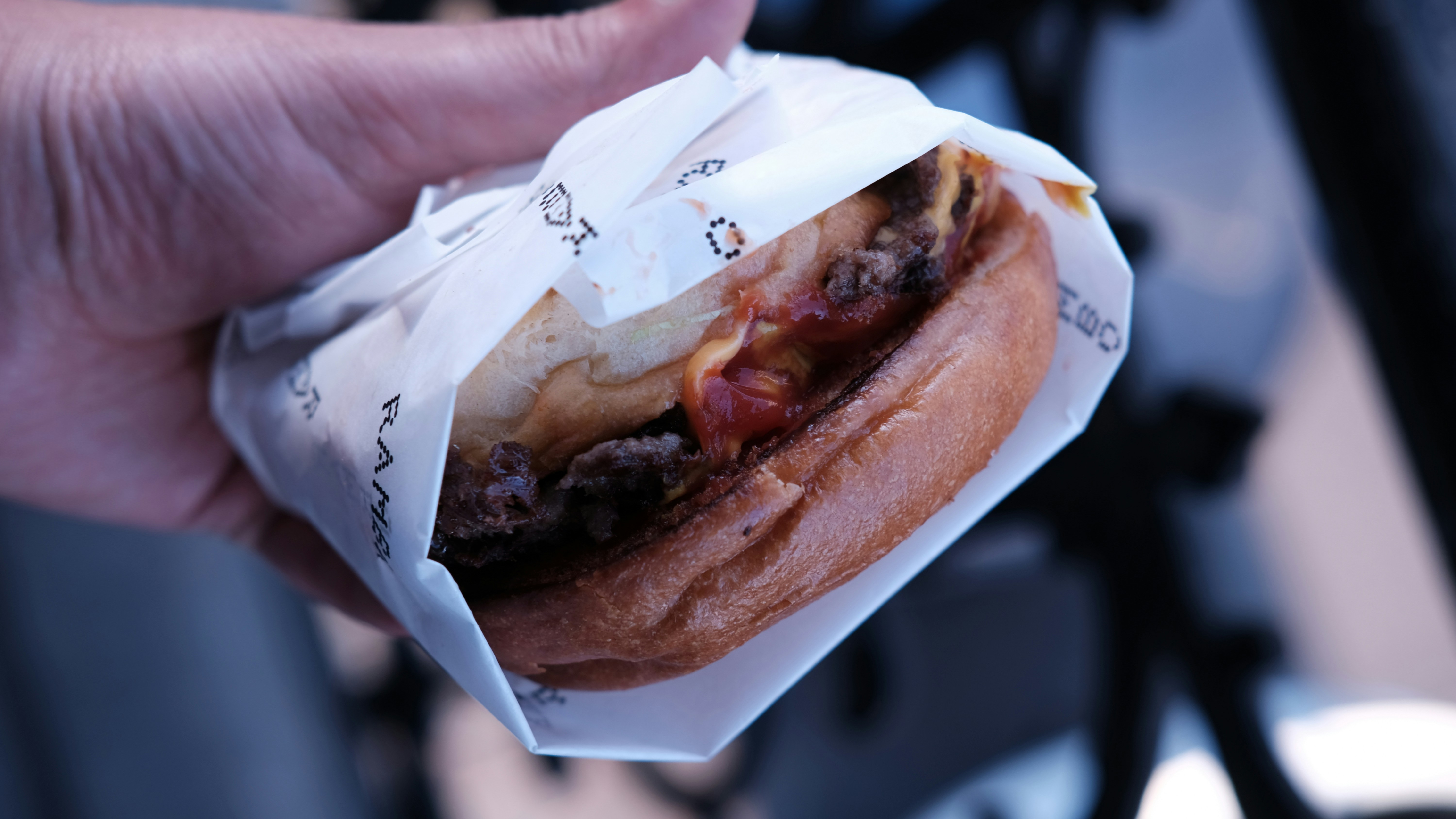 A hand holding a gourmet burger wrapped in paper, showcasing its juicy layers and toppings against a blurred background.