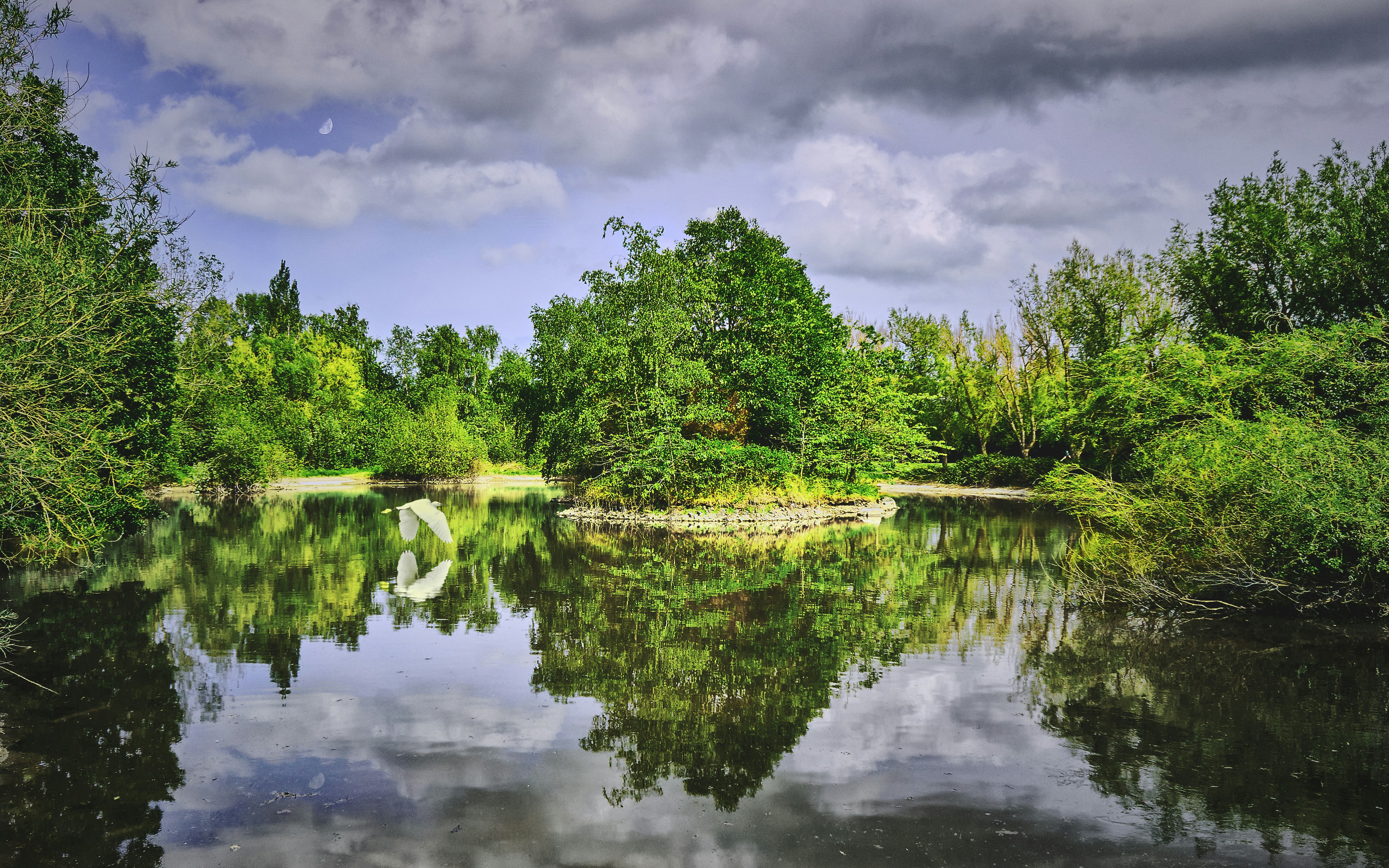 Lush greenery surrounds a calm pond, with a small island at its center reflecting the vibrant foliage and cloudy sky above.