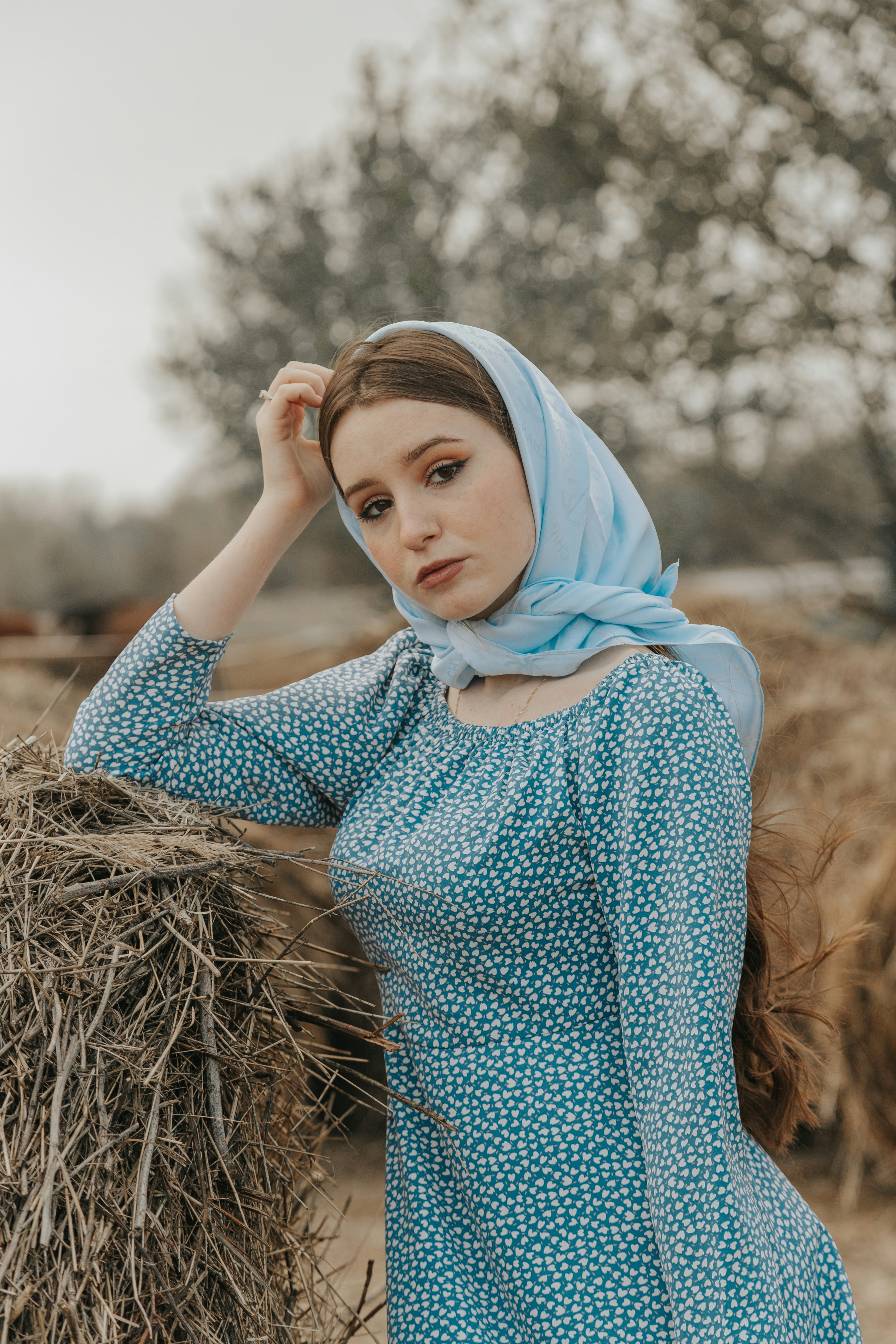 A young woman in a blue dress and light blue scarf poses thoughtfully beside a haystack, embodying a blend of cultural heritage and contemporary style.