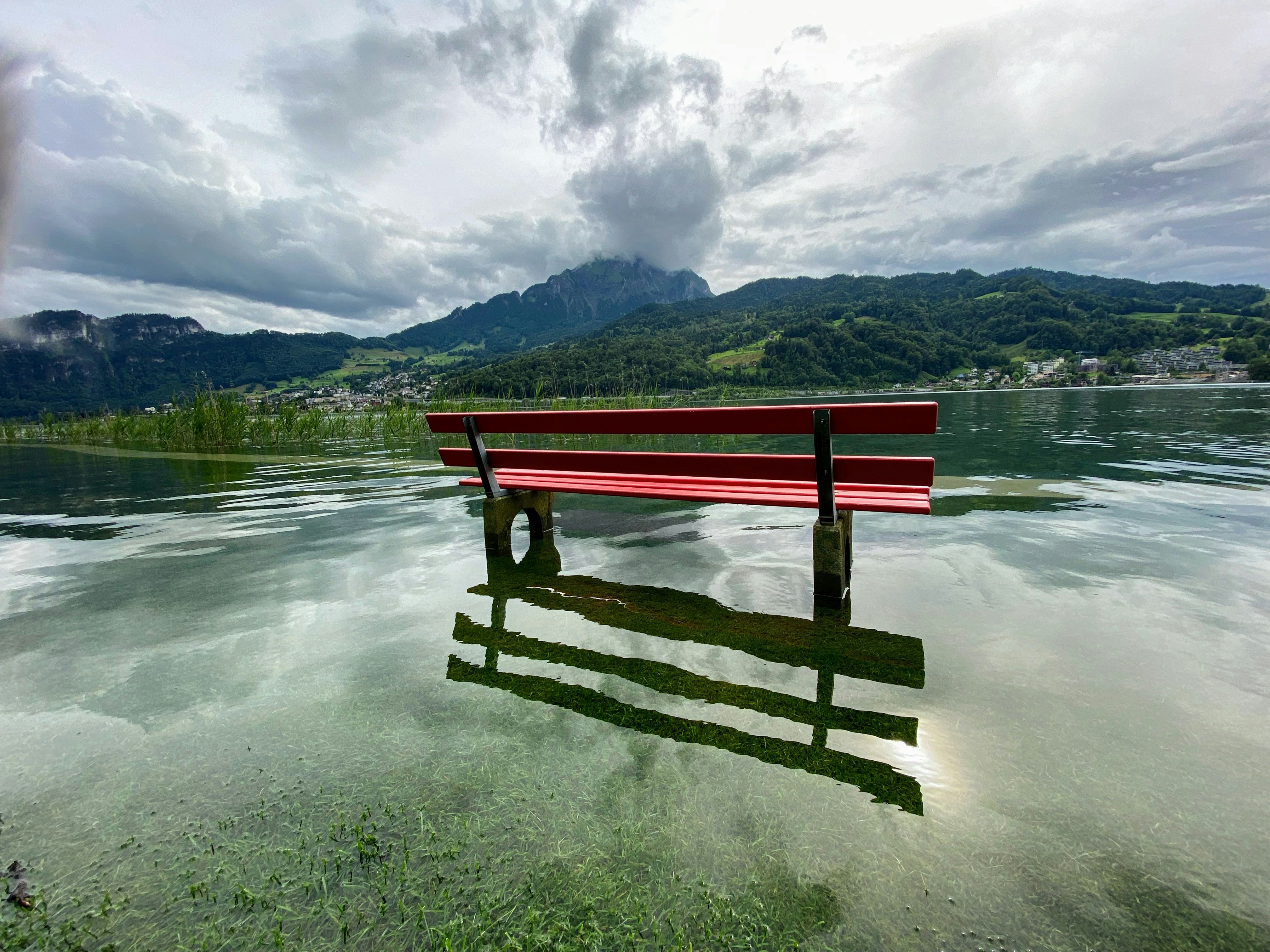 A surreal and quiet scene captures the impact of a flood at Lake Lucerne (Vierwaldstättersee) in Switzerland. A vibrant red wooden bench stands partially submerged, its legs disappearing into the clear, rising waters that have claimed the surrounding meadow. Below the surface, the green grass remains visible, creating a hauntingly beautiful underwater landscape. In the background, the jagged peaks of Mount Pilatus are draped in heavy, dramatic clouds, framing the Swiss Alps in a moody, overcast light. This powerful image blends serene beauty with the stark reality of natural forces.