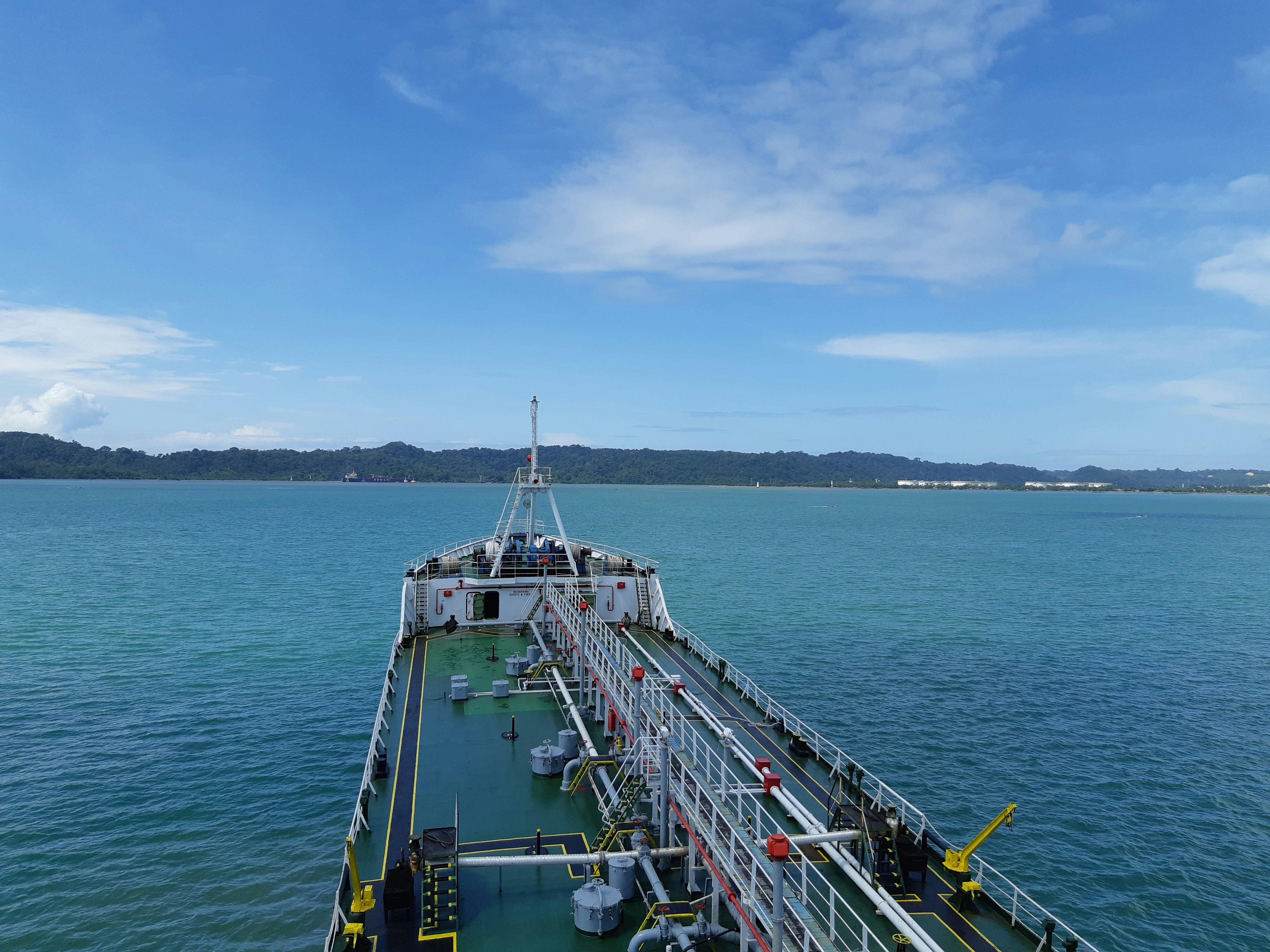 Wide view of a ship deck extending toward a turquoise sea with a distant shoreline. The scene emphasizes the vessel's structural railings and walkway against a clear sky.