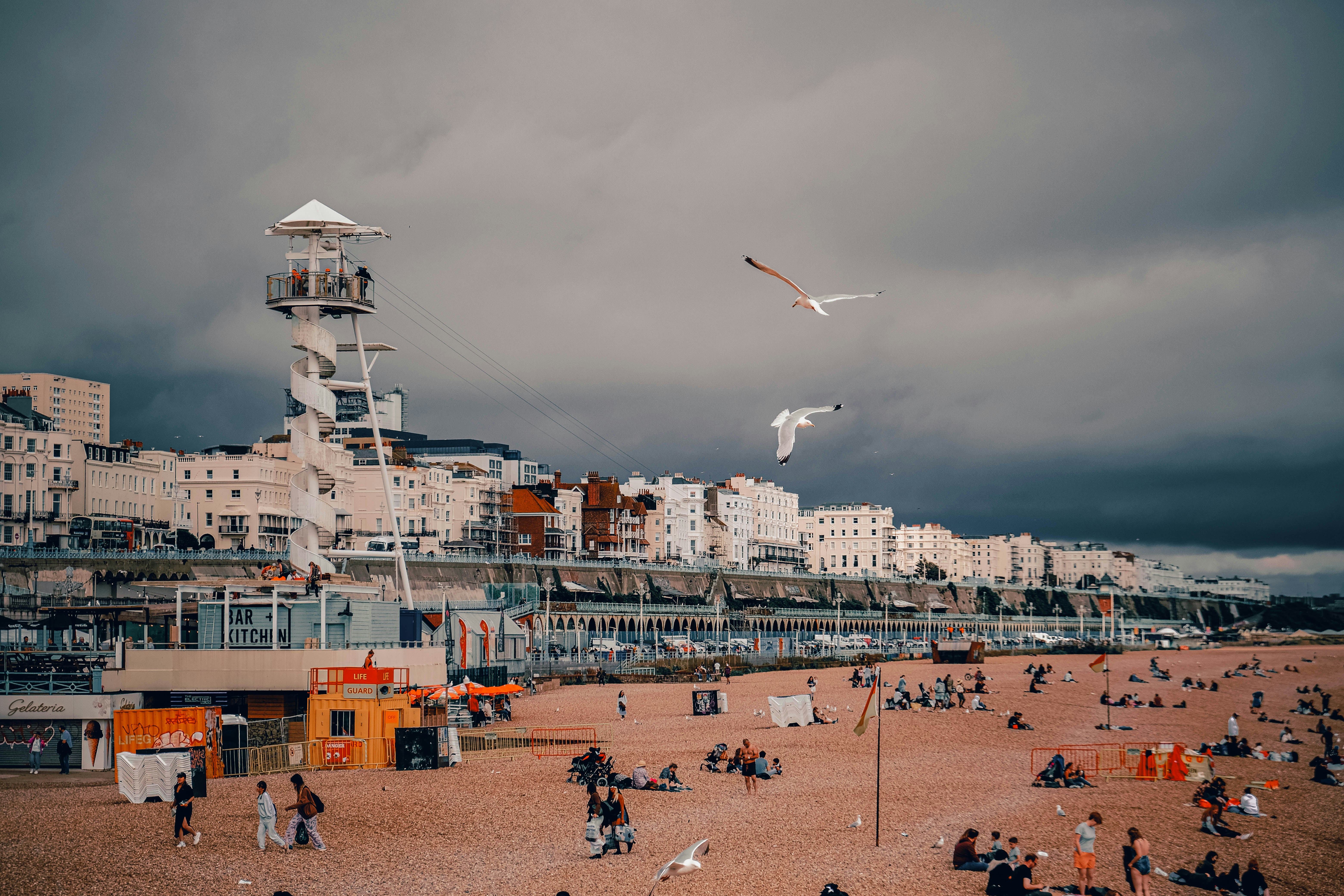 Lifeguard tower stands watch over a bustling beach filled with visitors, as seagulls soar under a moody sky. 