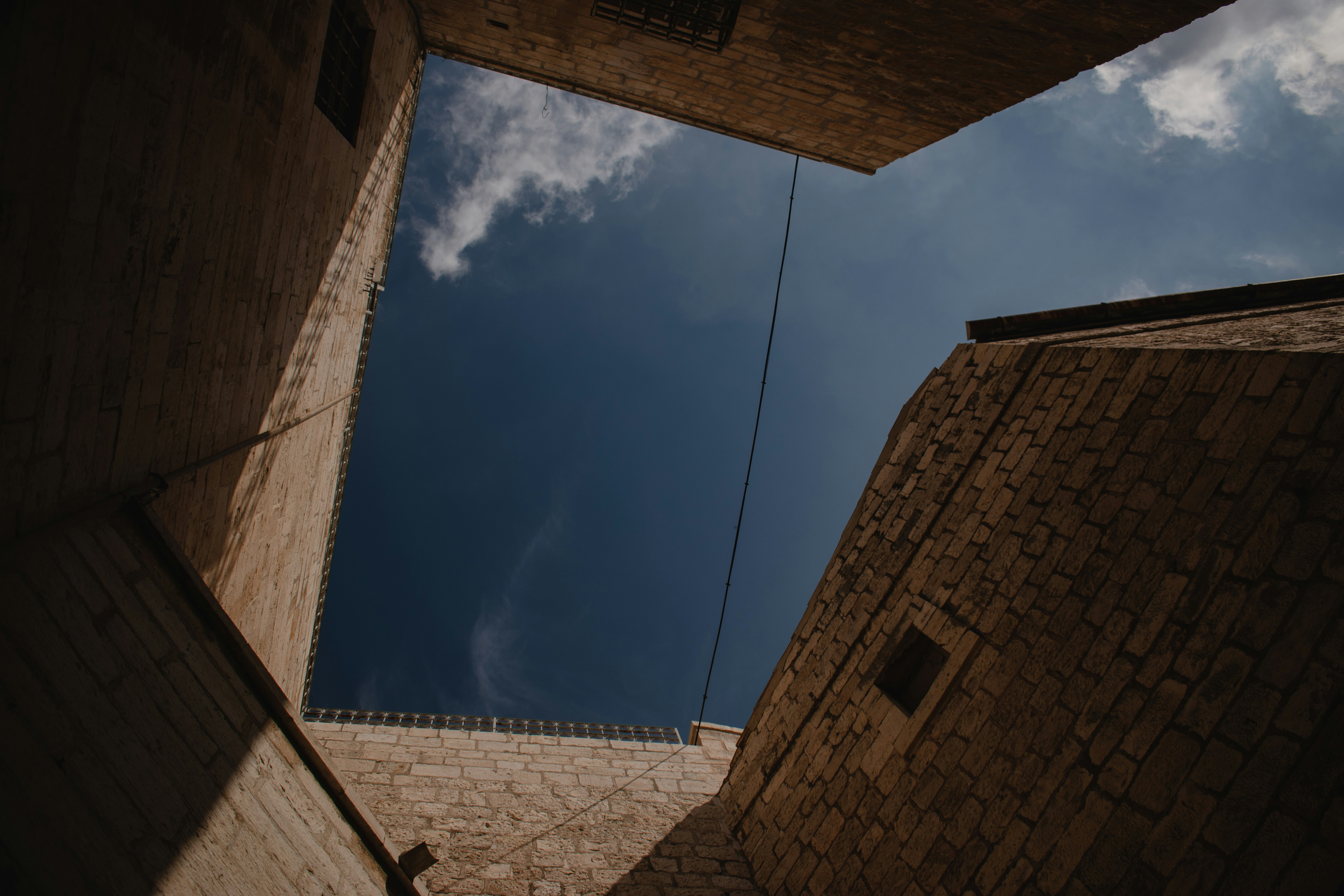 View of a narrow courtyard framed by stone walls, showcasing a vibrant sky above with scattered clouds. The composition emphasizes the architectural lines converging towards the sky.