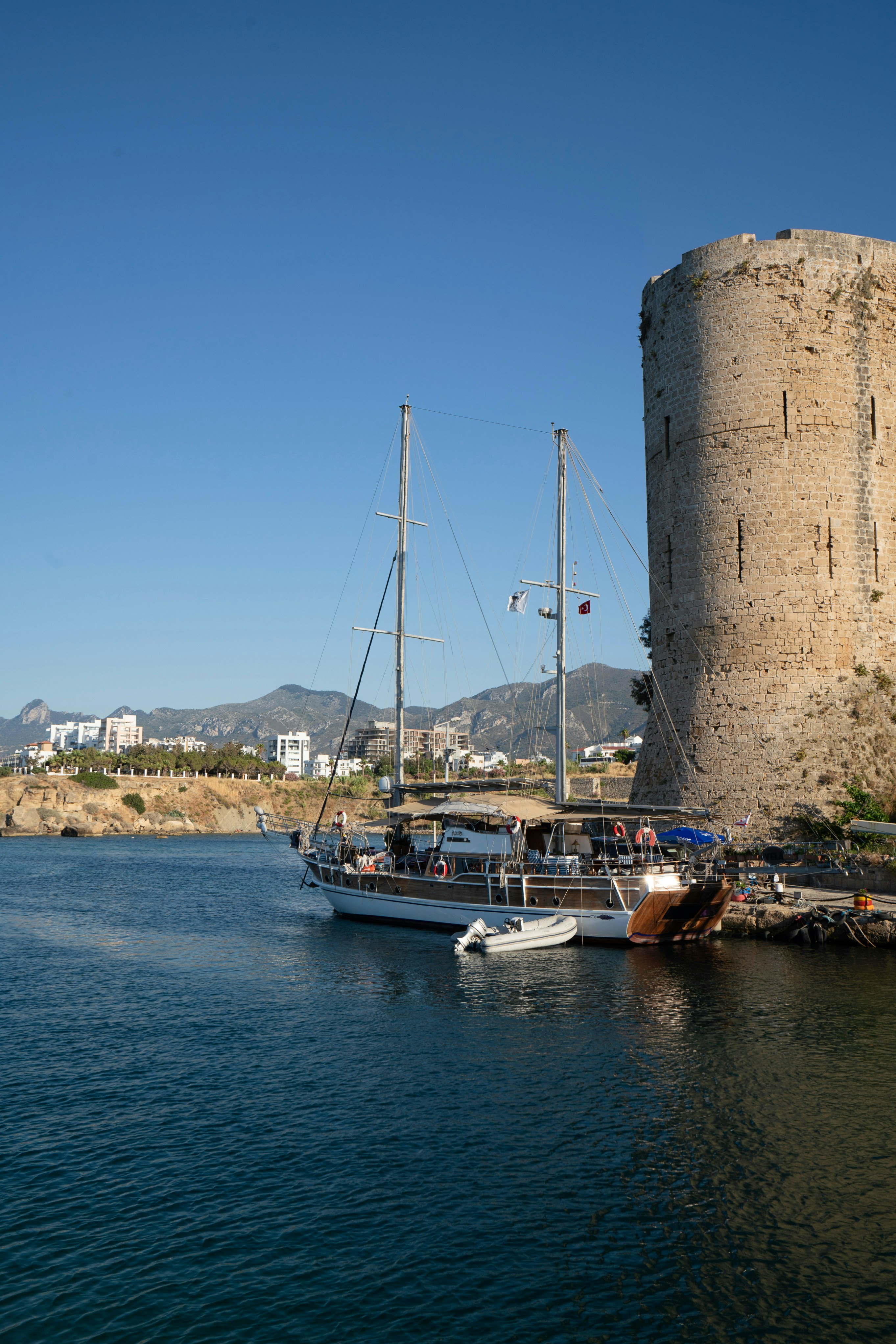 Sailing vessel docked beside an ancient stone tower, with a backdrop of mountains and modern buildings under a clear blue sky.
