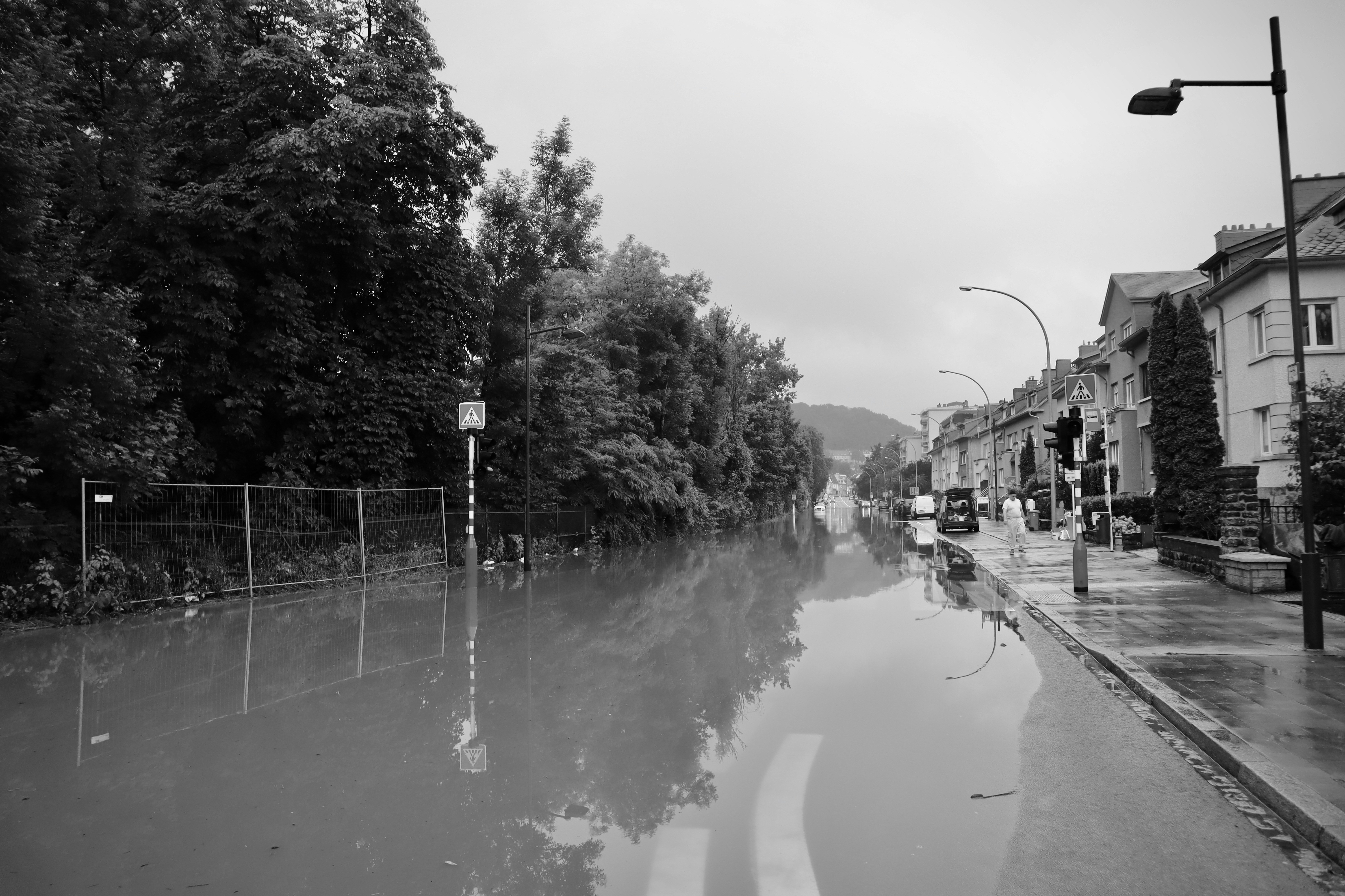 Flooded urban street reflecting surrounding buildings and trees under overcast sky.
