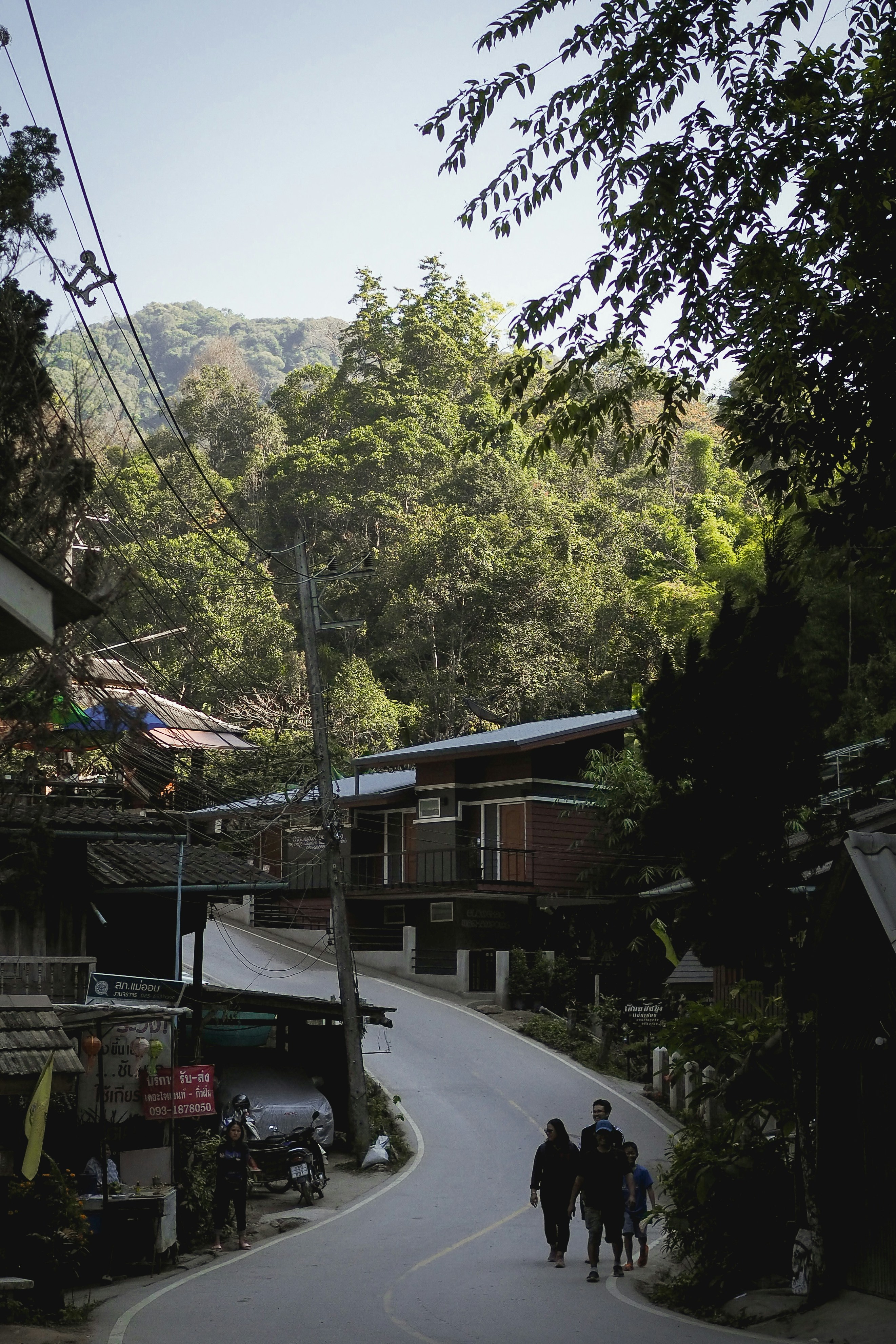 Two figures walking along a winding road lined with rustic buildings and lush greenery. The scene captures the serene atmosphere of a hillside village.