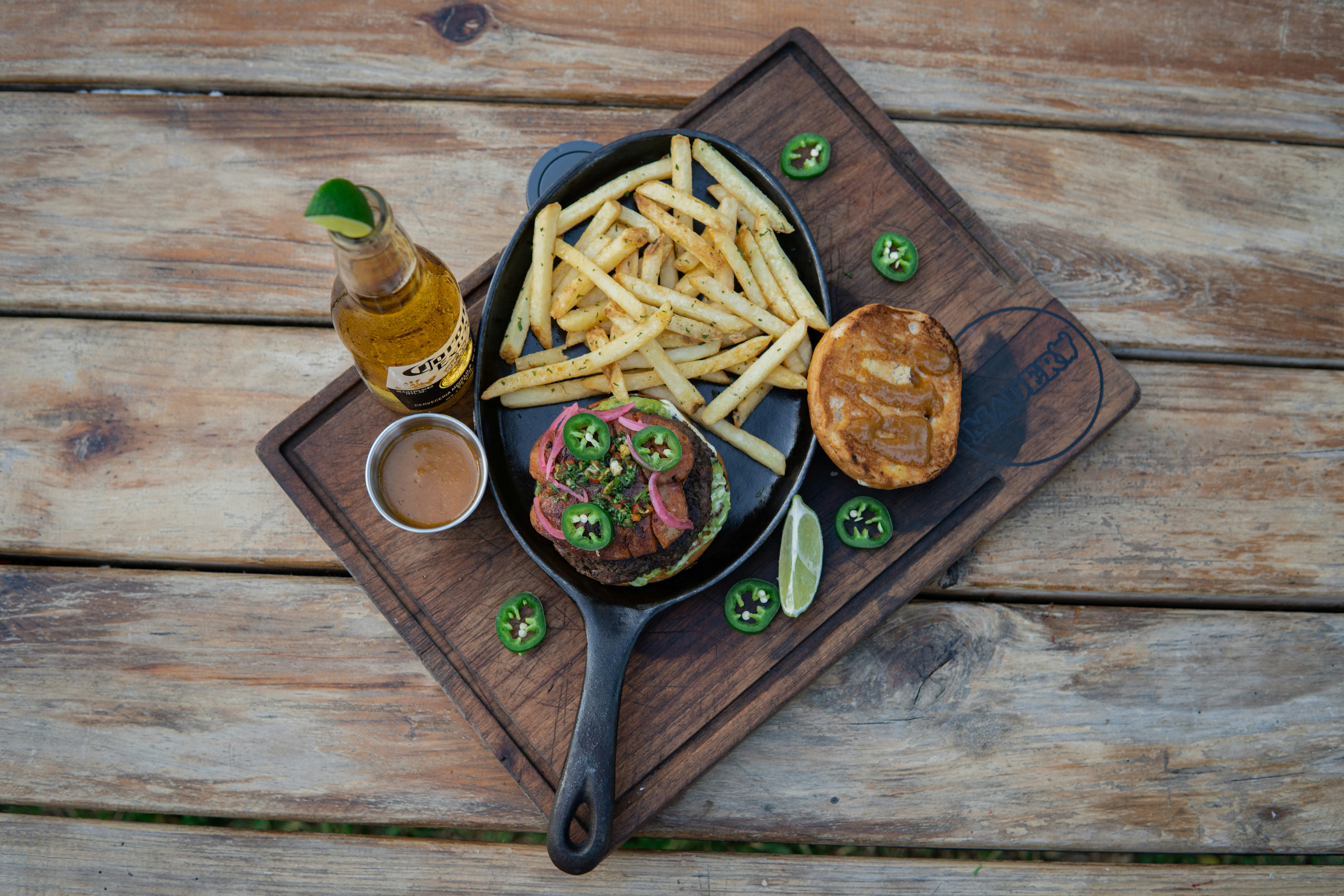A gourmet burger served in a cast iron skillet alongside crispy fries, jalapeños, and a refreshing drink, all set on a wooden table.