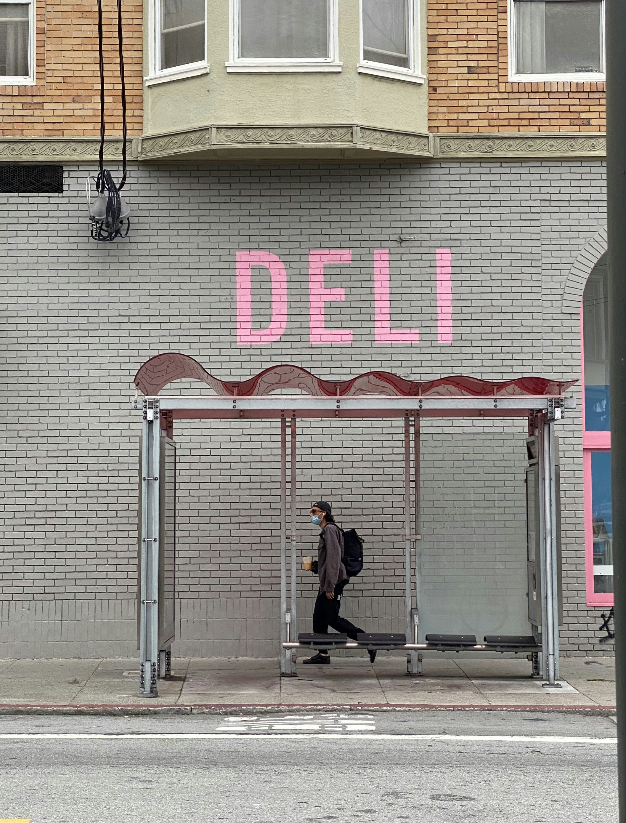 A pedestrian walks past a deli with a prominent pink sign, showcasing the vibrant life of the city. The scene captures the essence of urban living.