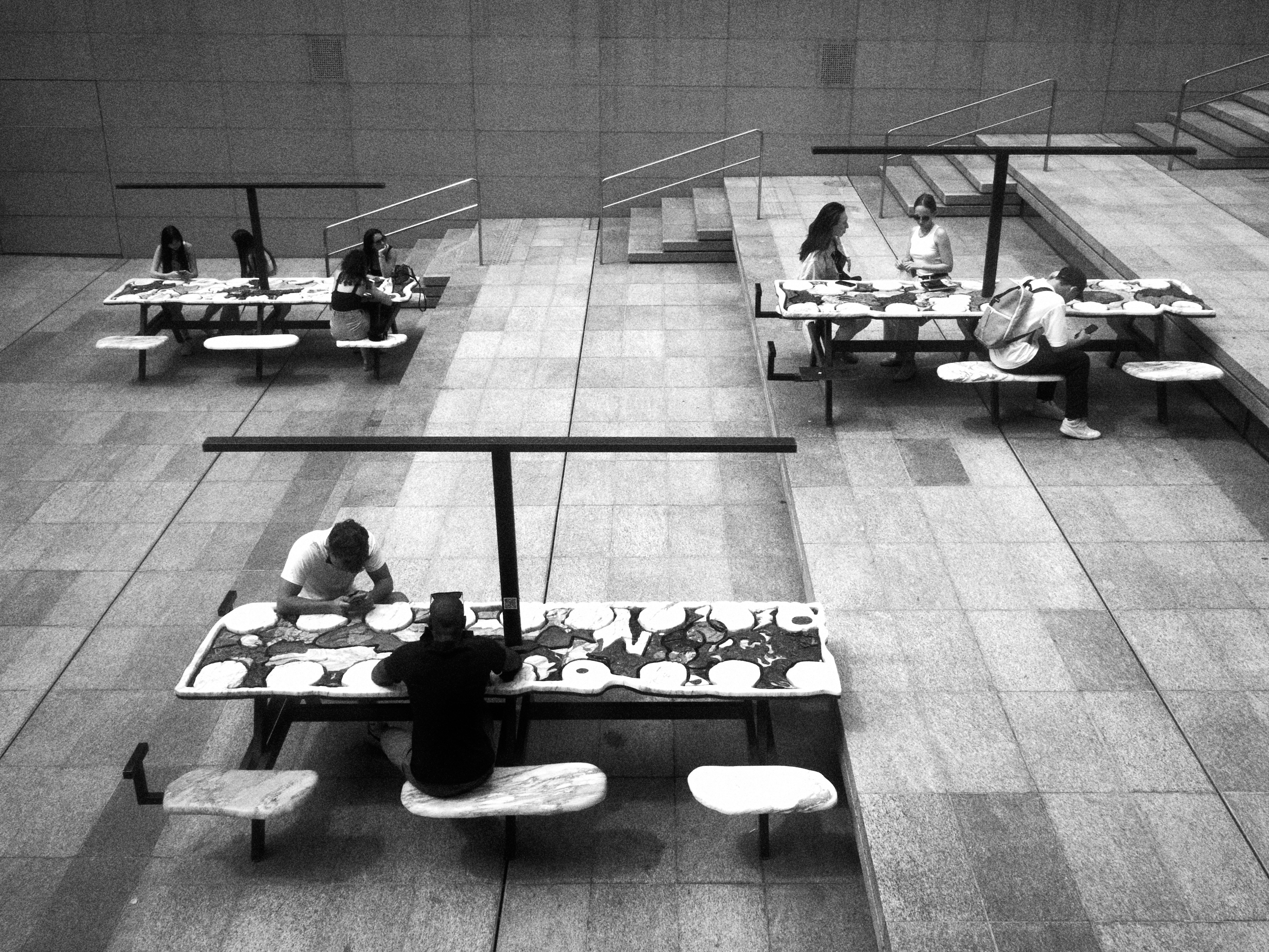 Monochrome scene of several groups seated at long tables in an urban courtyard, captured from above; emphasis on communal dining and architectural rhythm. (This is a photograph, not a painting or illustration.)