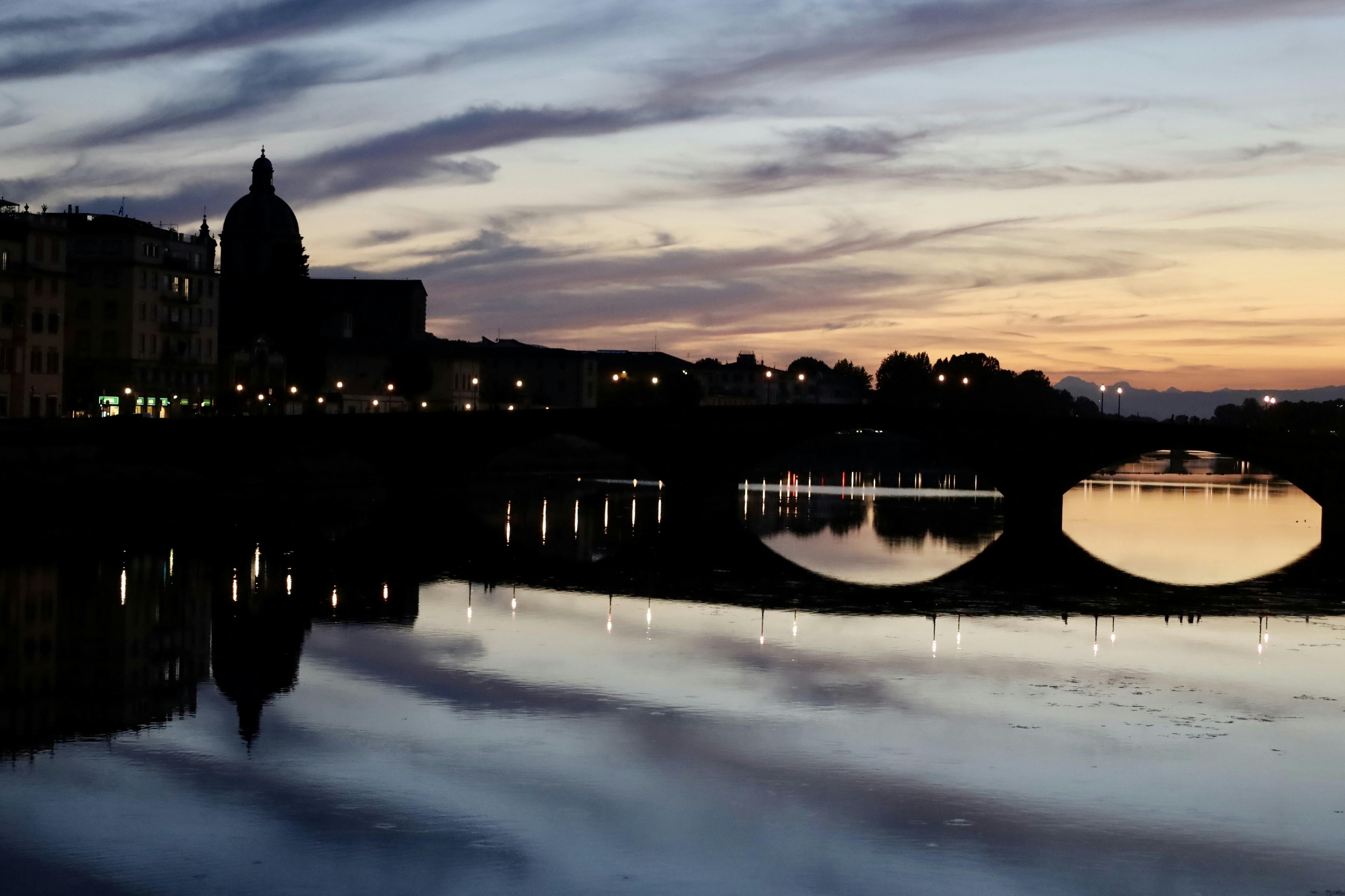 Silhouette of Florence buildings and bridge reflecting in the Arno River at sunset.