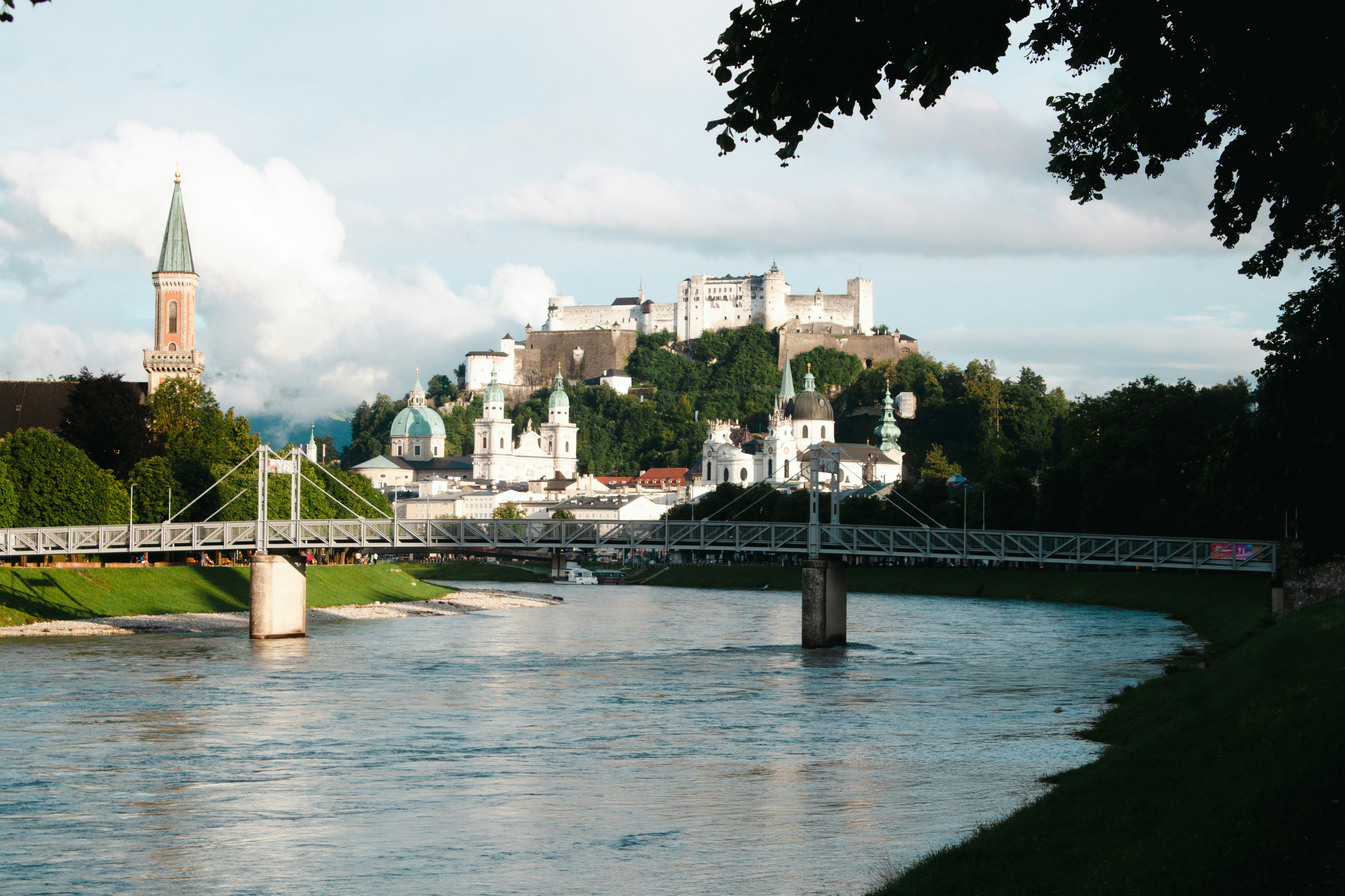 Bridge crossing a serene river with a historic fortress perched on a hill under a partly cloudy sky.