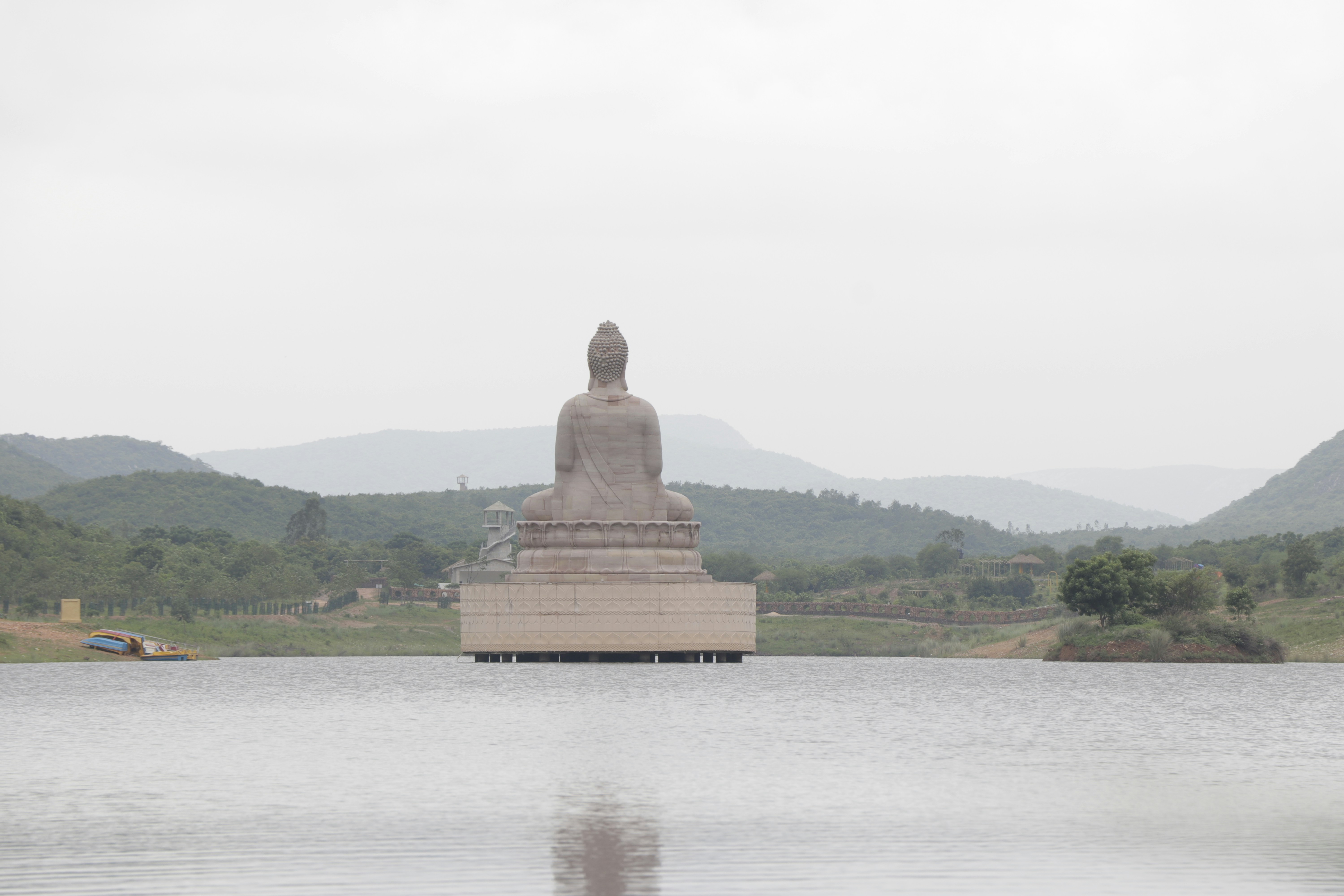 Majestic statue of a seated Buddha overlooking a serene lake, surrounded by lush hills under a soft, overcast sky.