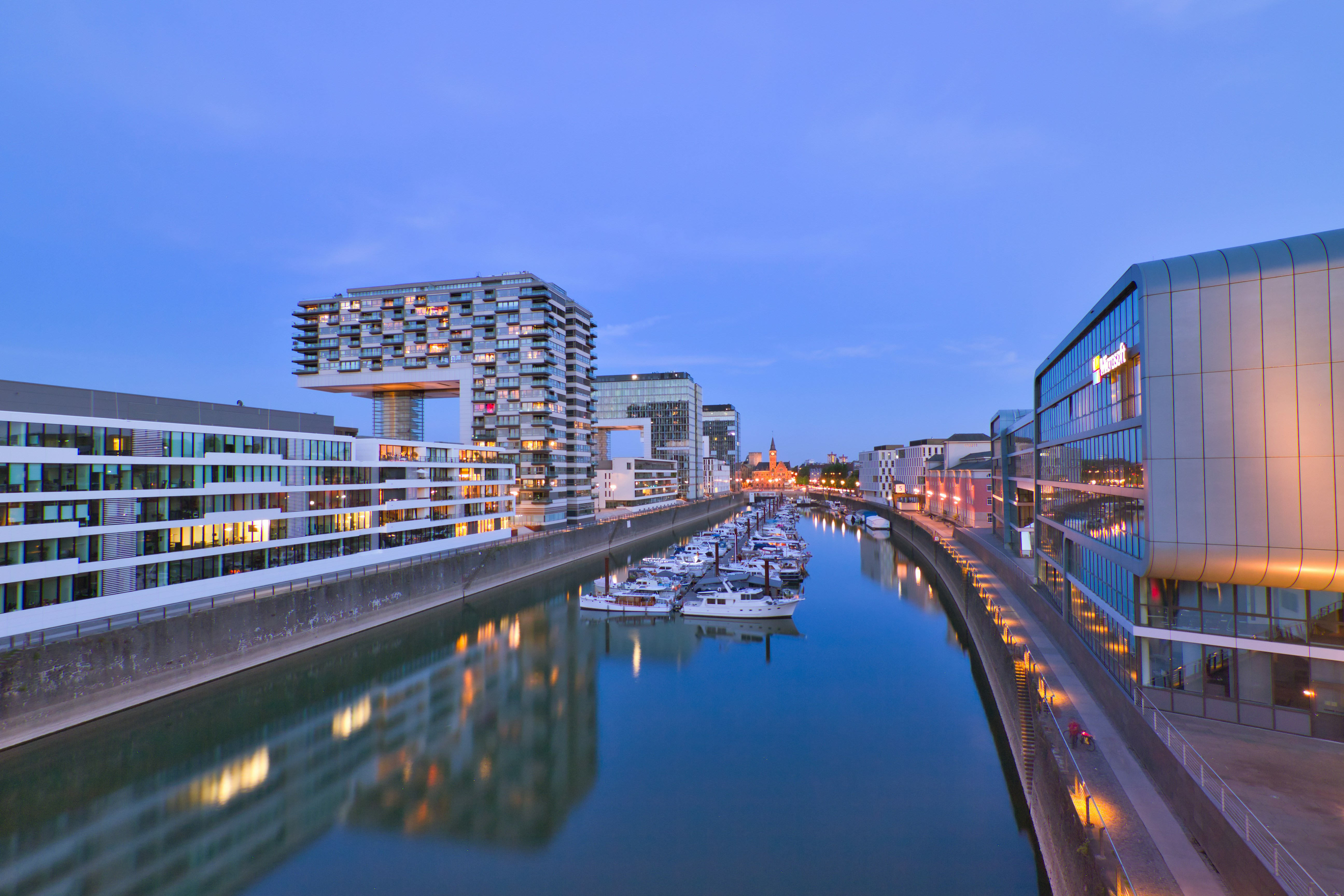 Contemporary buildings line a tranquil canal at dusk, reflecting in the calm water under a blue sky.