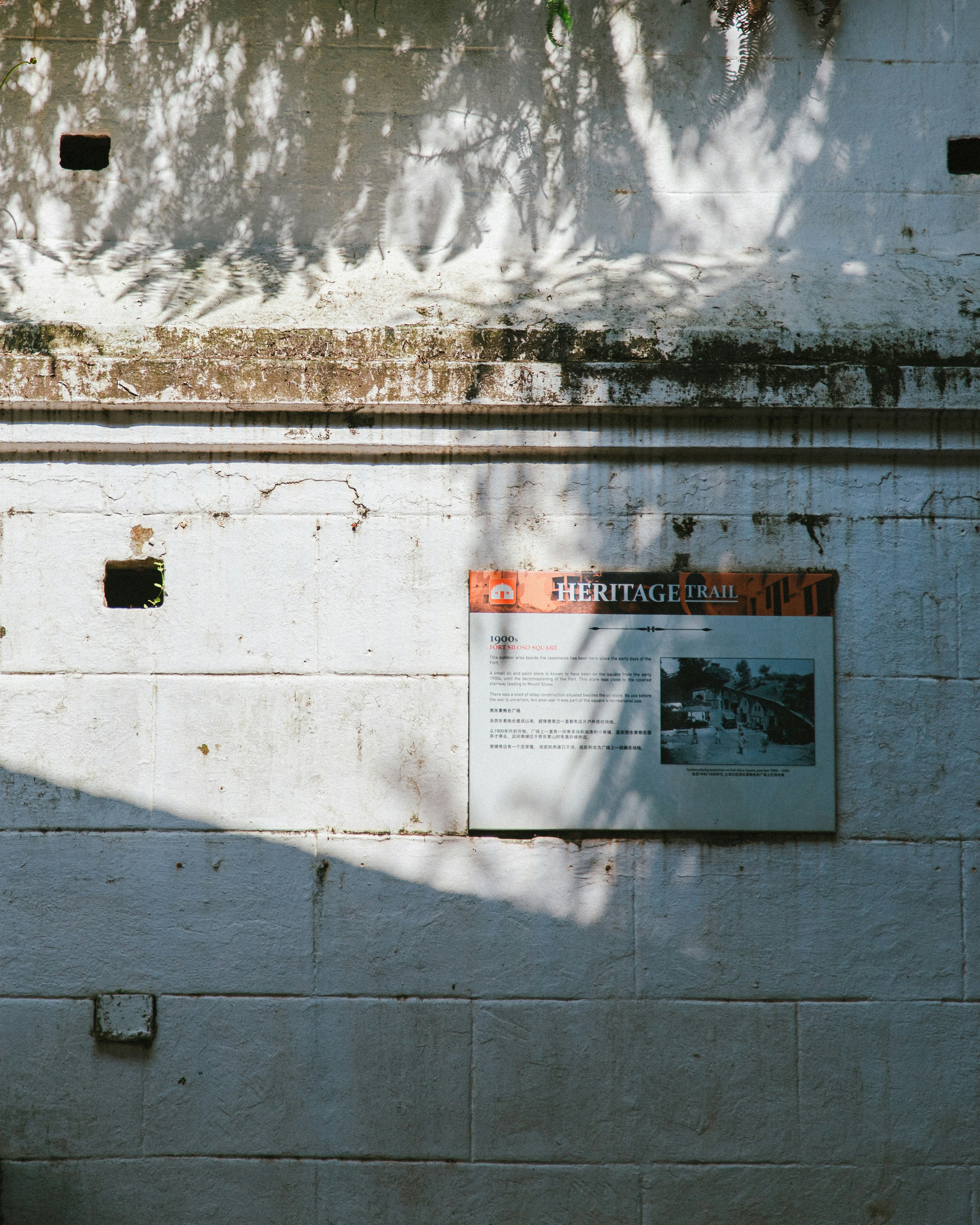Heritage trail sign on a textured stone wall with dappled sunlight and shadow patterns.