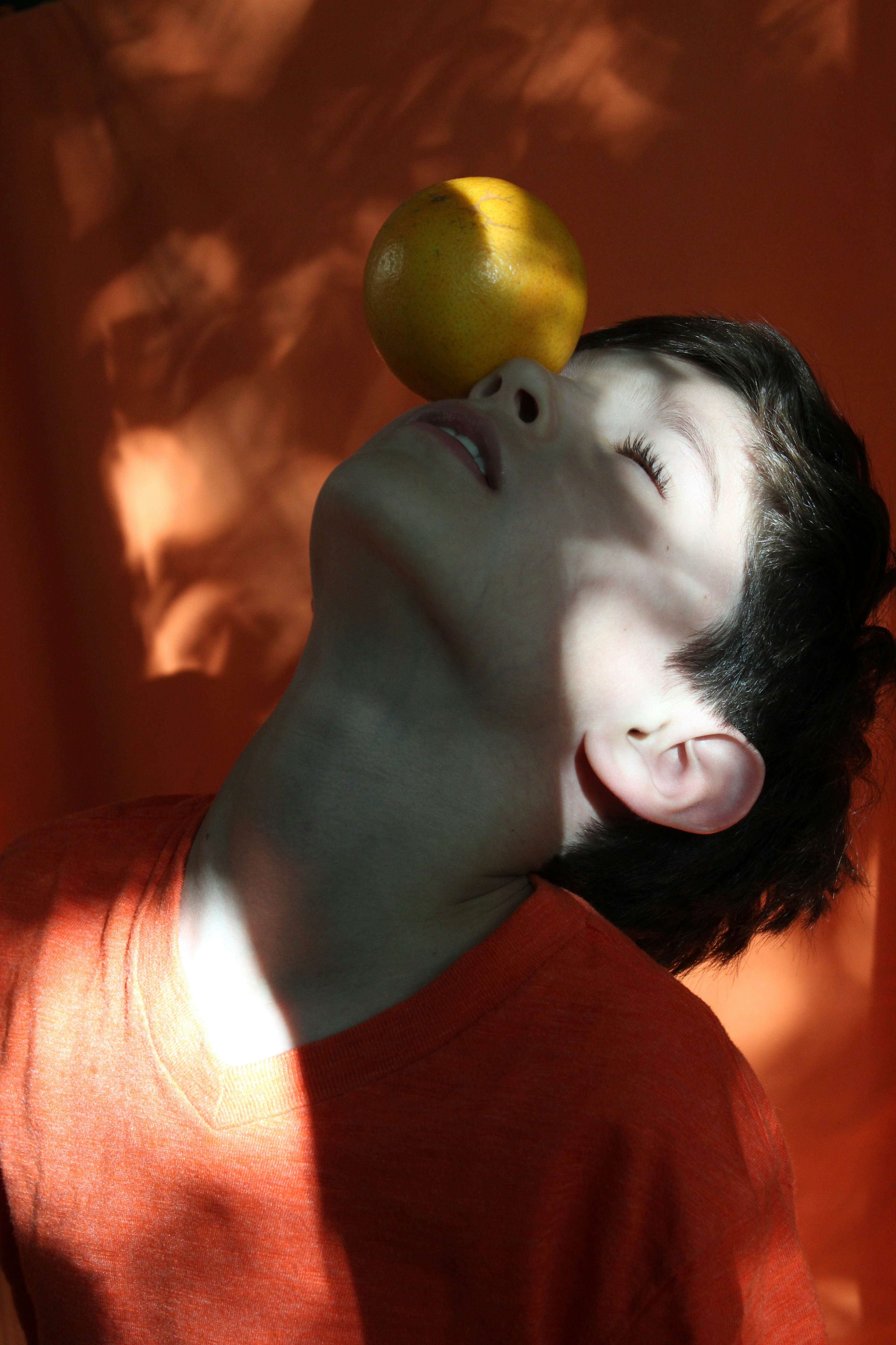 Child balancing an orange on their face against an orange backdrop with dappled sunlight.