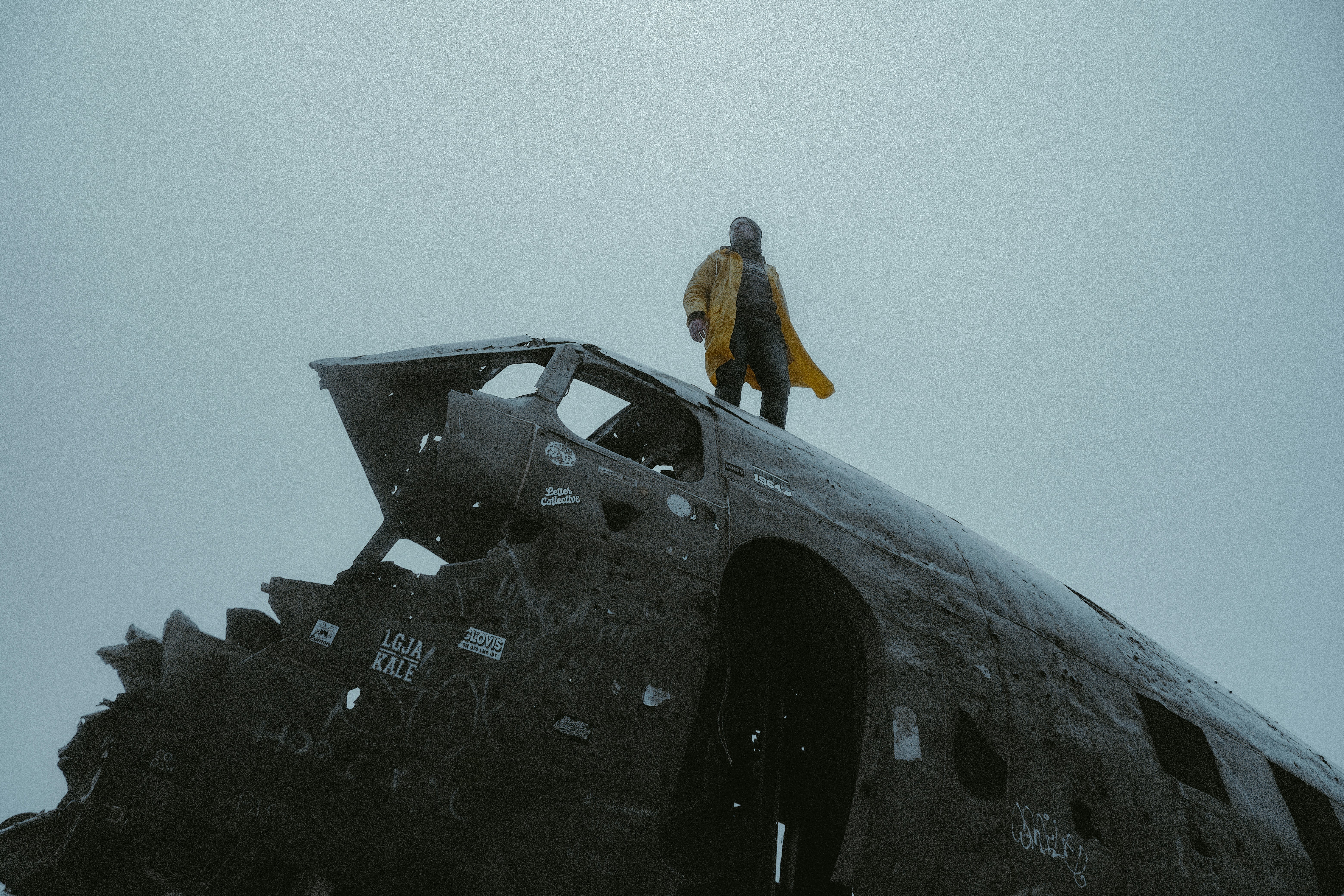 A figure in a yellow raincoat stands atop a derelict airplane fuselage, surrounded by a bleak, overcast sky. The scene evokes themes of desolation and exploration.