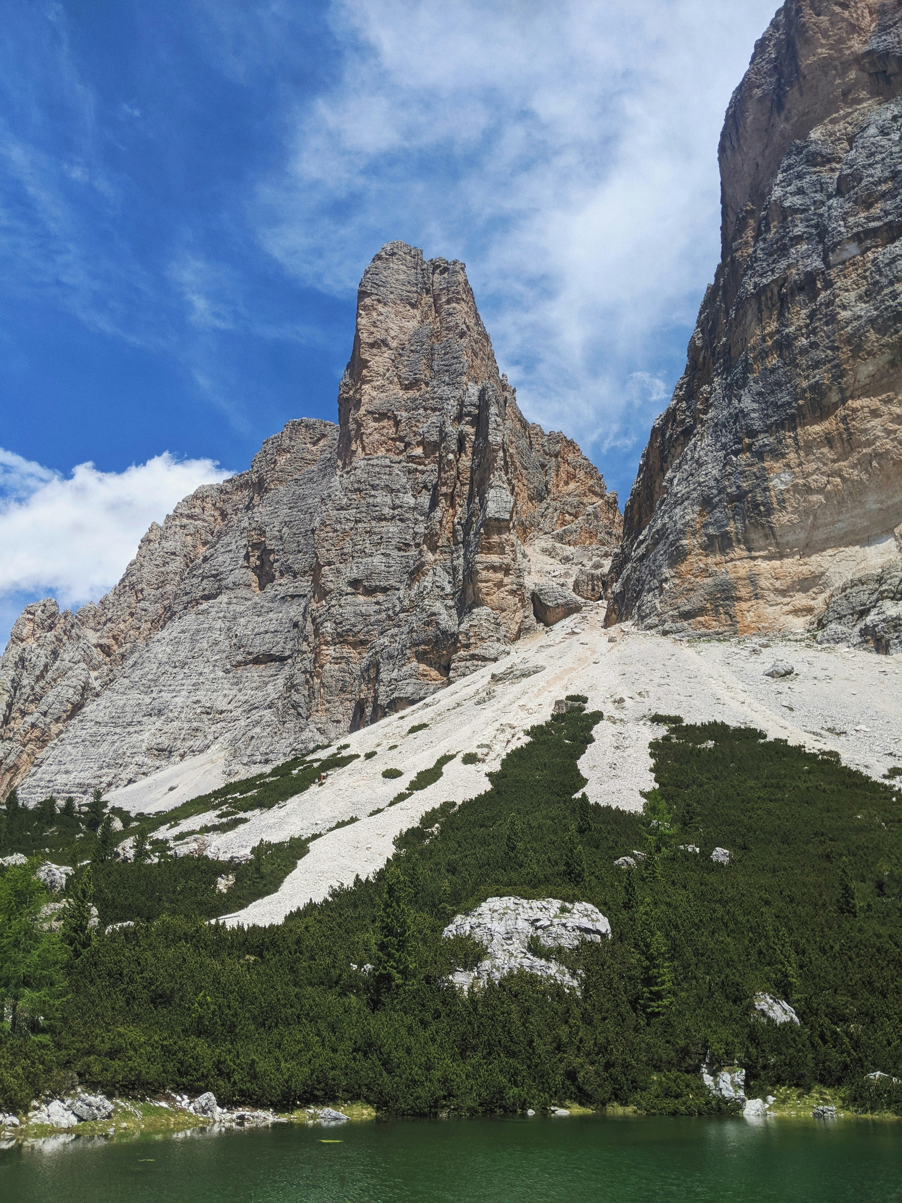 Towering rocky cliffs rise above a tranquil green lake, framed by lush vegetation under a bright blue sky.