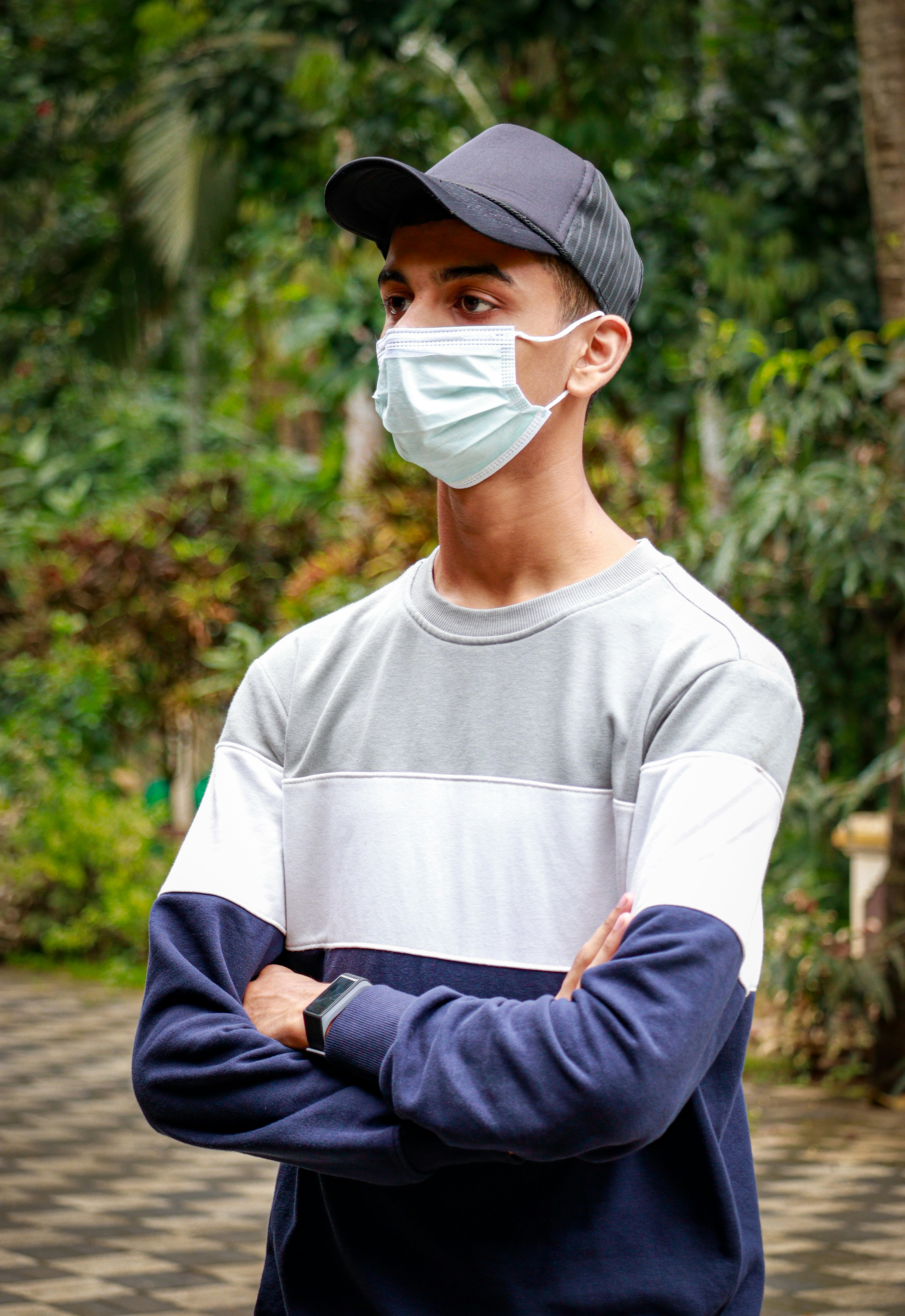Young man wearing a mask and cap stands with arms crossed in a lush outdoor setting, conveying a sense of introspection.