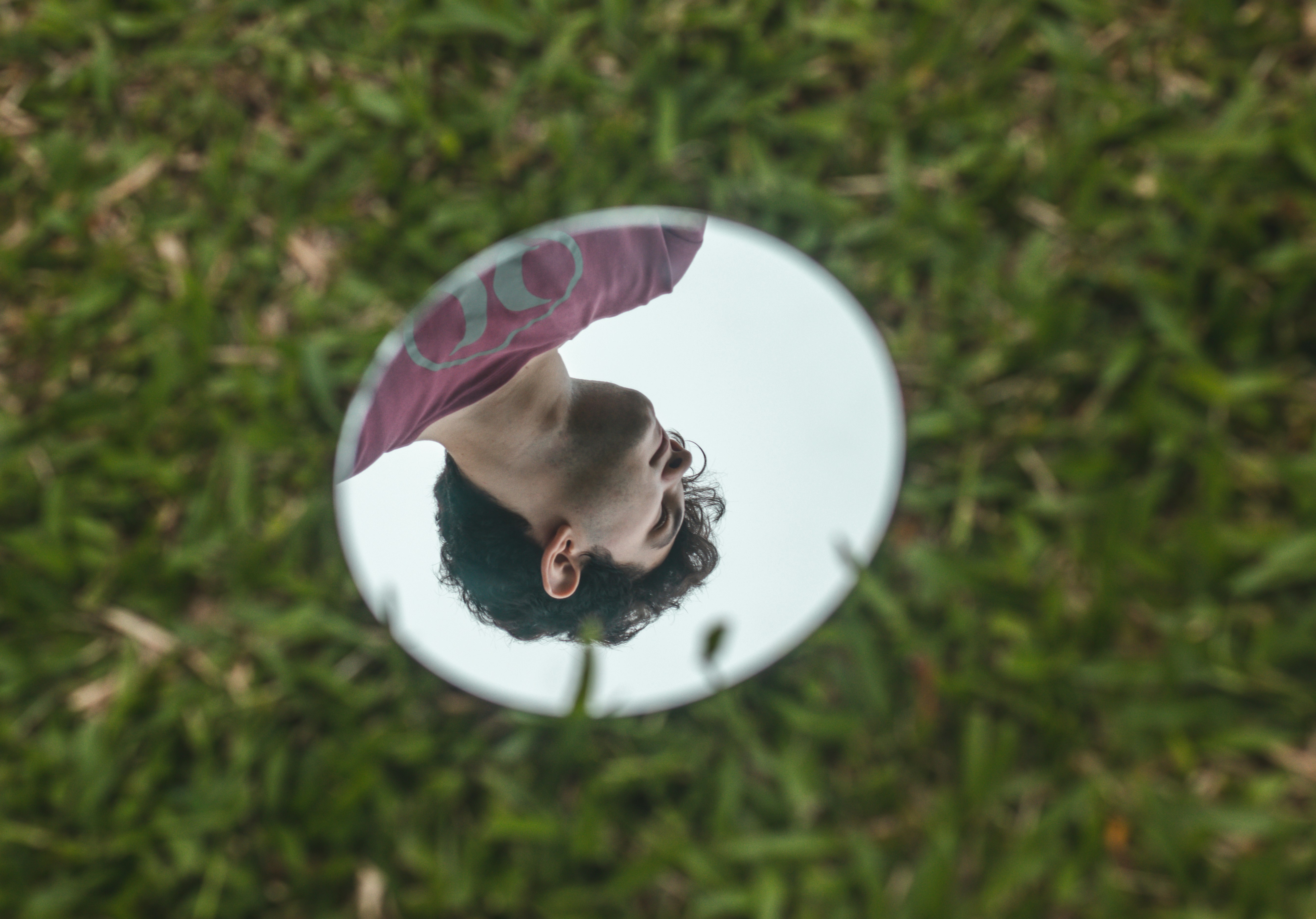 A reflection of a young man captured in a circular mirror resting on lush green grass, creating a unique perspective on identity and nature.