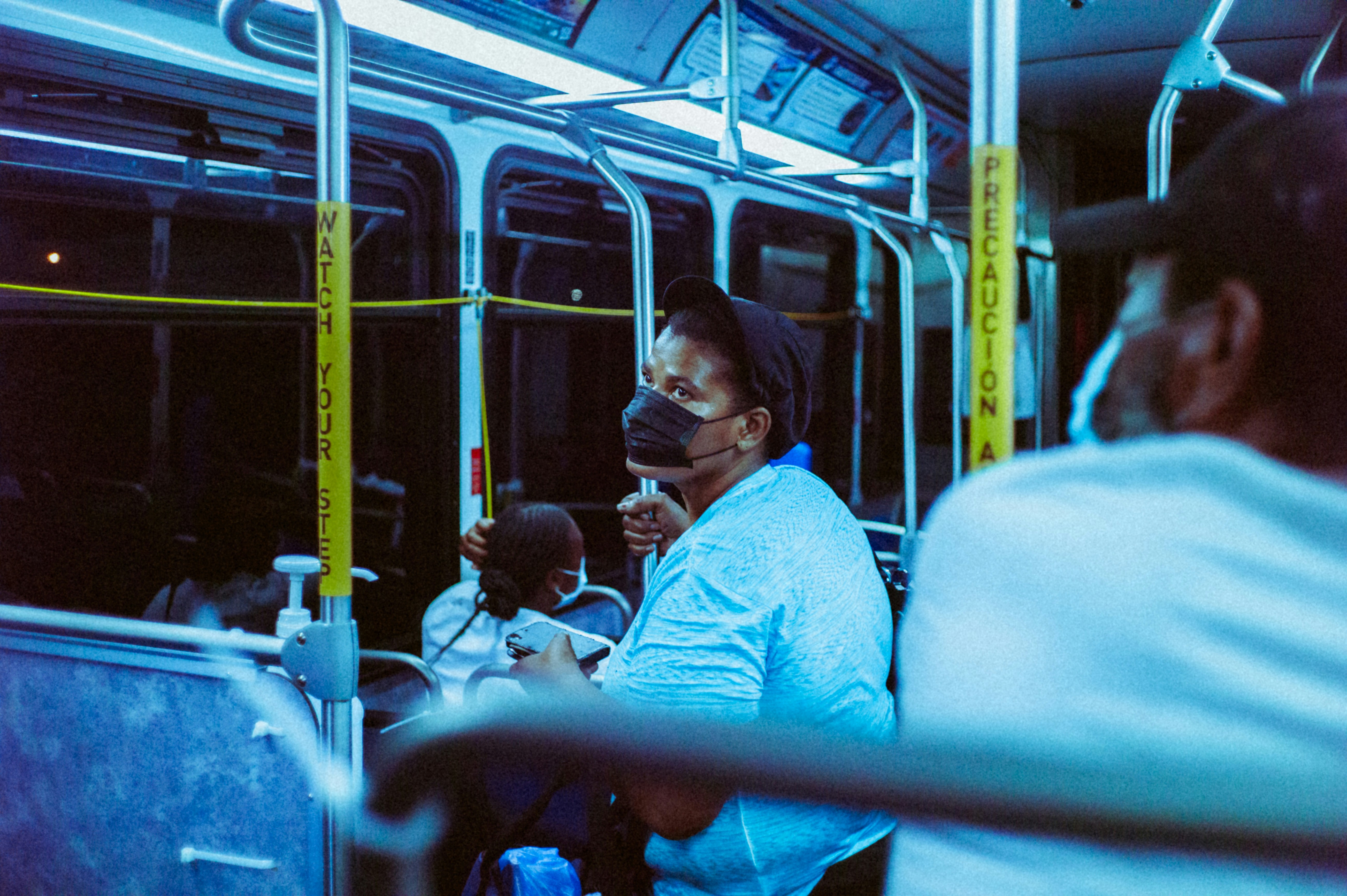Candid photo of a masked passenger in a blue shirt standing inside a dimly lit bus, bathed in cool blue interior lighting.