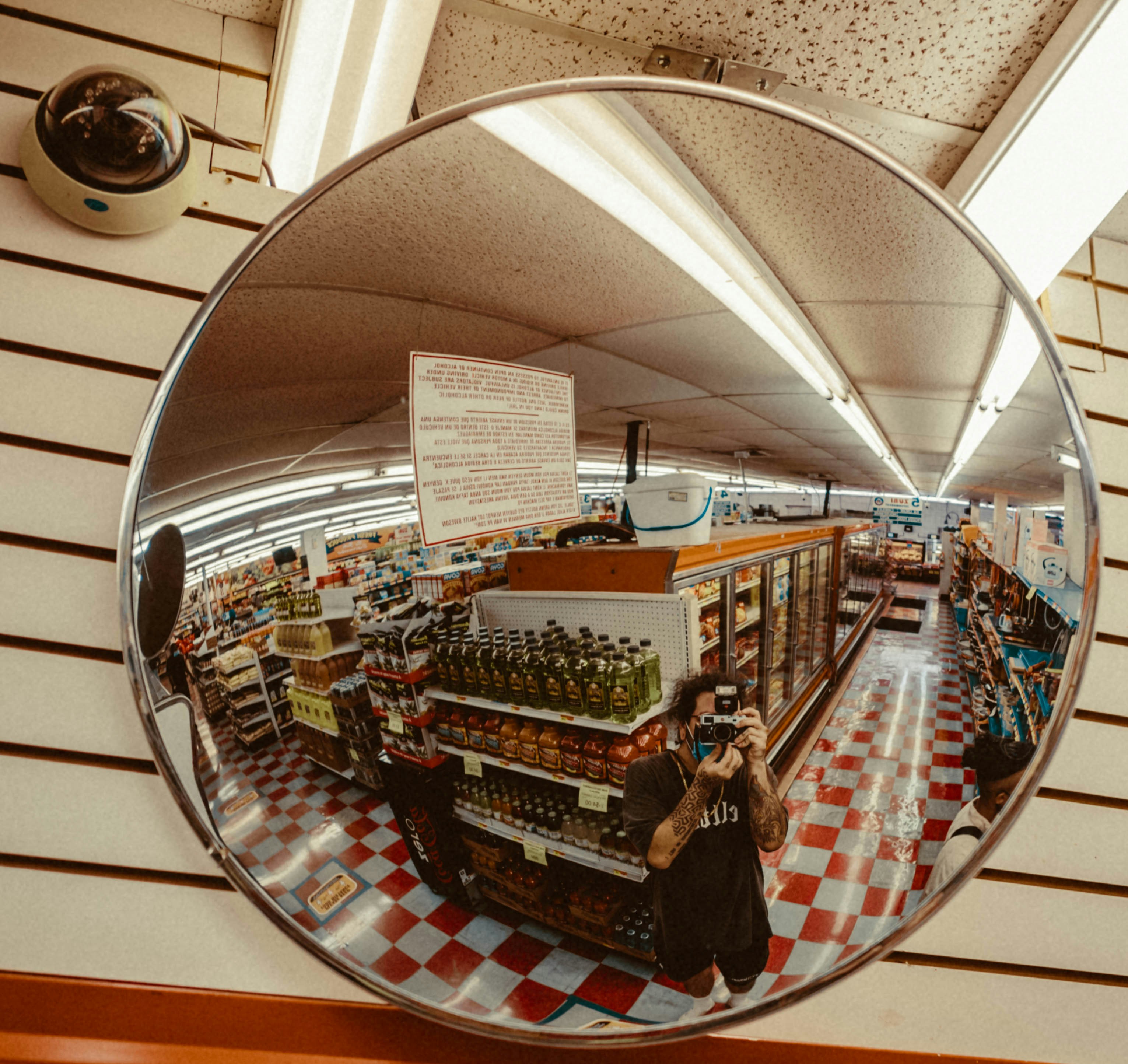 Circular mirror reflecting a grocery store aisle with colorful products and a person taking a photo. The scene captures the vibrant atmosphere of a local shop.