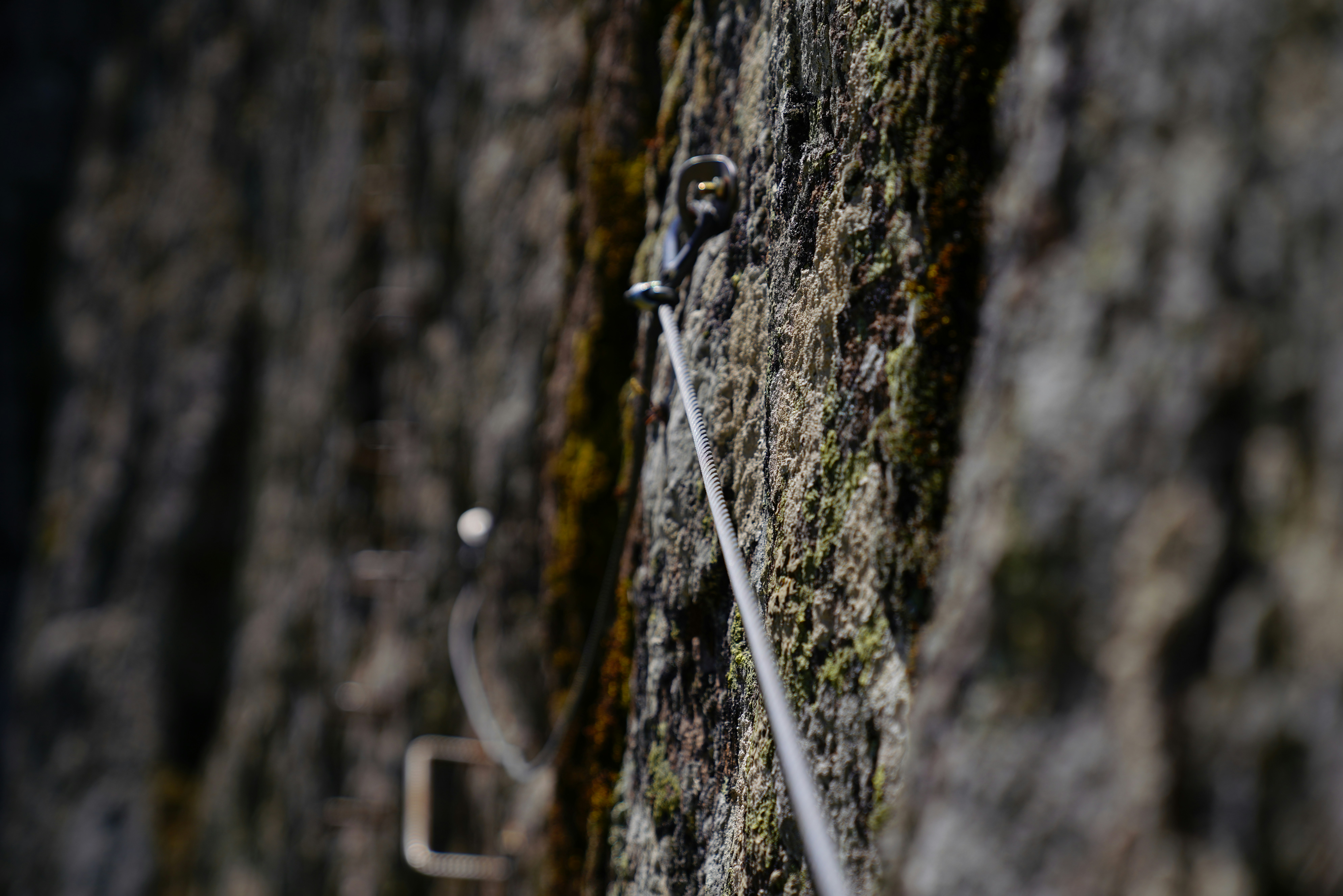 Metal climbing gear secured to a rugged rock face, showcasing the texture and detail of the stone. Moss and weathering add character to the natural backdrop.