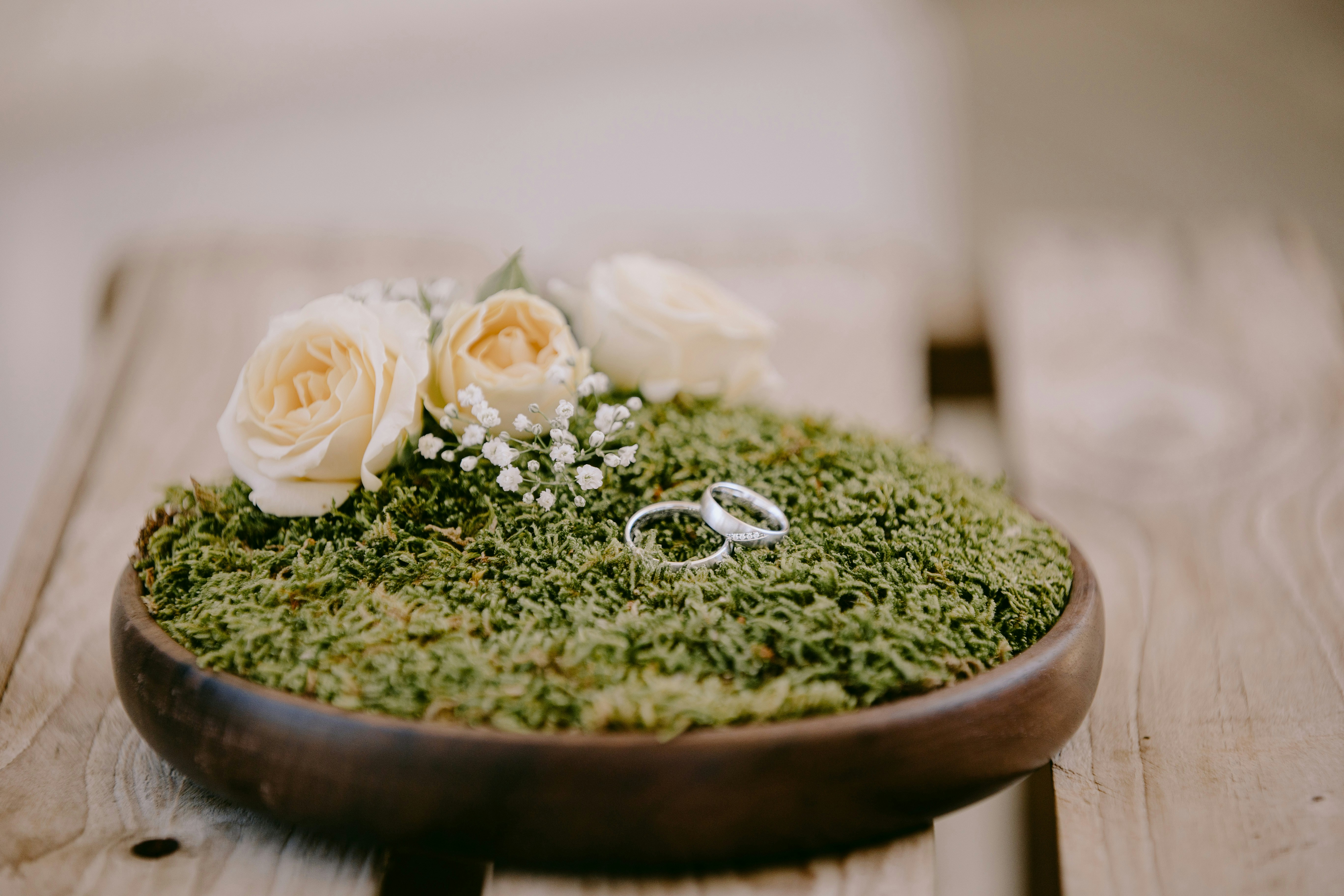 Delicate cream roses and baby's breath arranged on lush moss, featuring two silver wedding bands nestled together. 