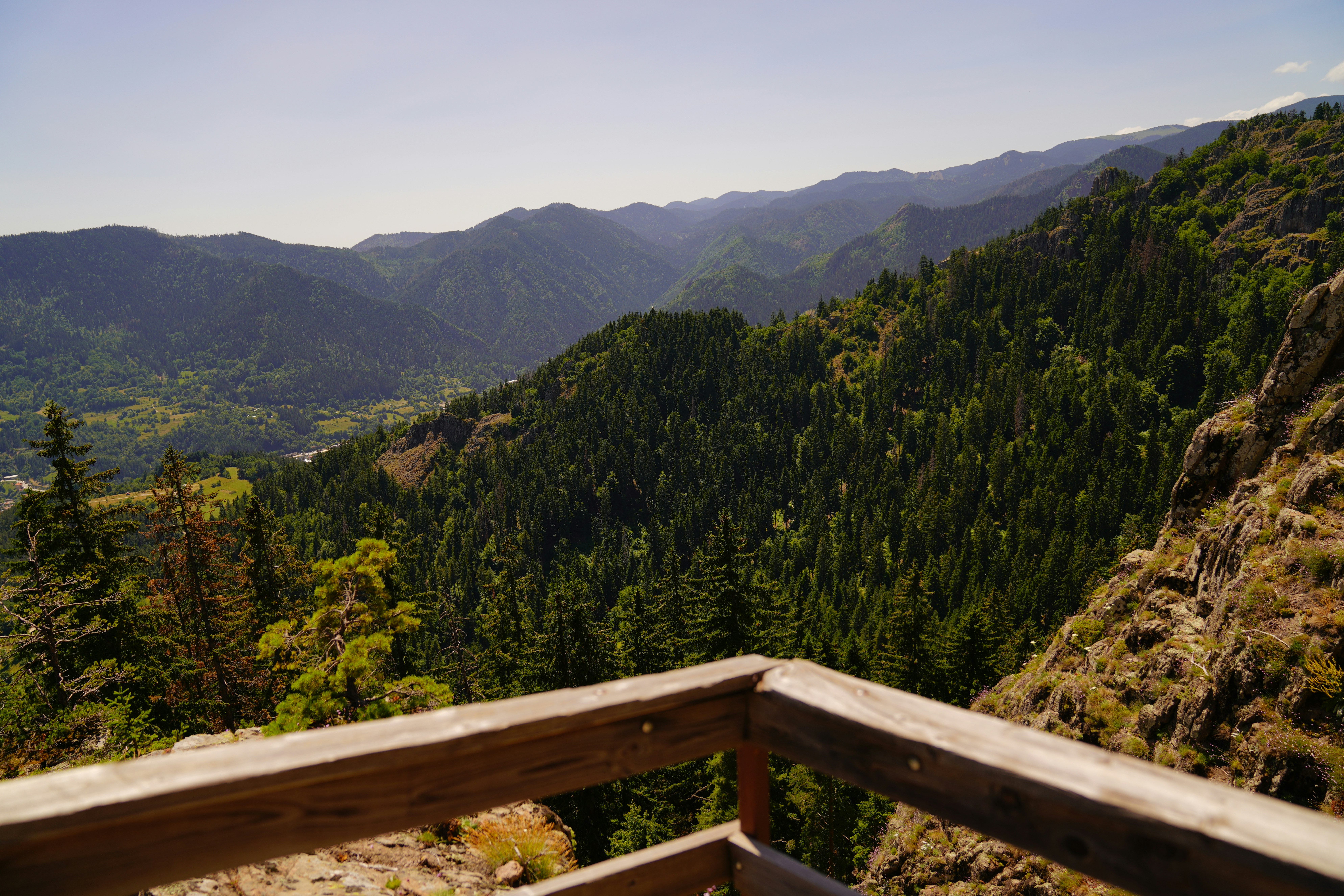Lush green mountains stretch into the distance under a clear blue sky, framed by a rustic wooden railing. The scene captures the serene beauty of nature from a high vantage point.