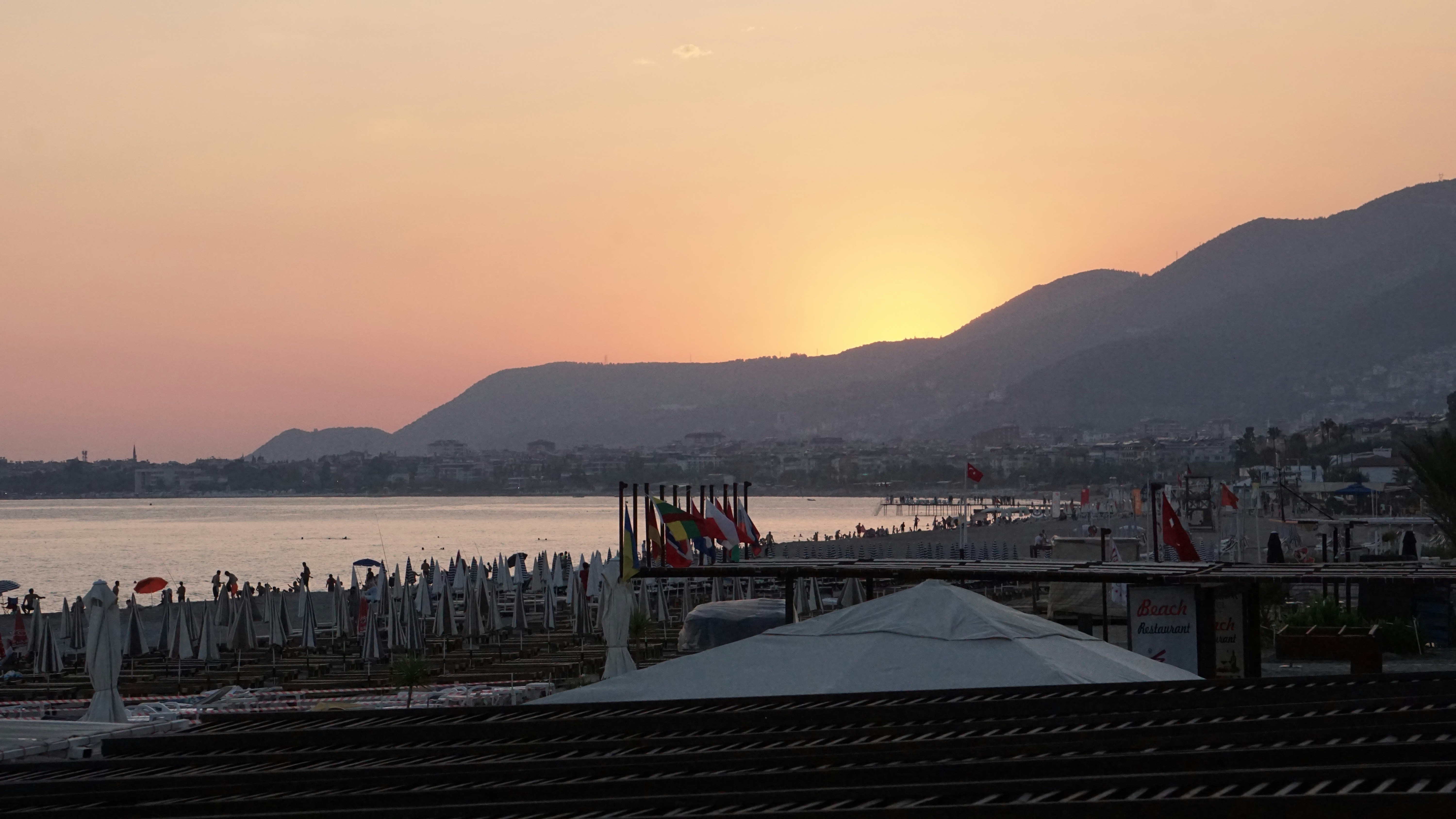 Golden sun dipping below the horizon, casting a warm glow over a tranquil beach lined with umbrellas and flags. 