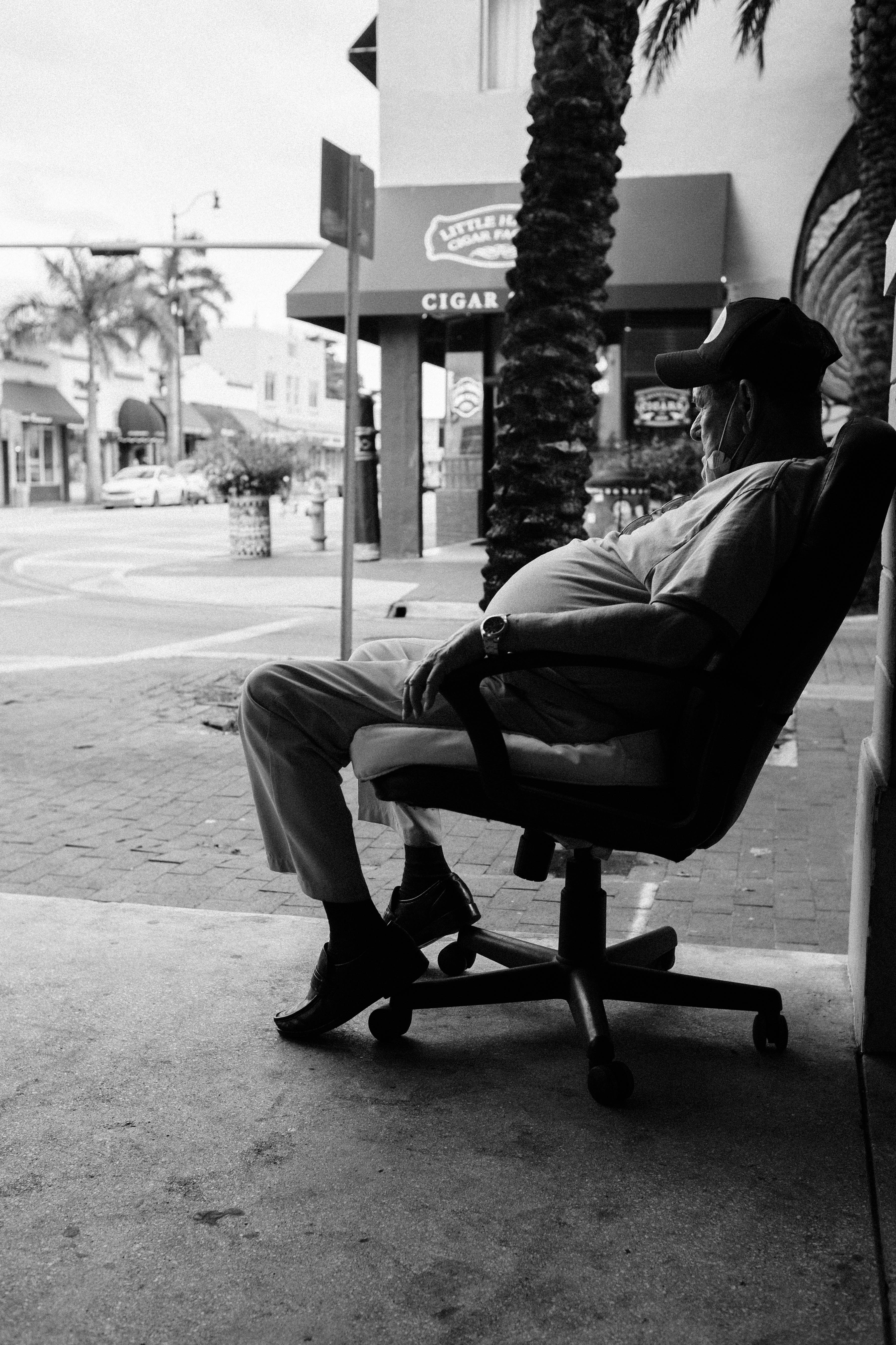 A man seated in a chair, lost in thought, against a backdrop of urban life, featuring palm trees and storefronts.
