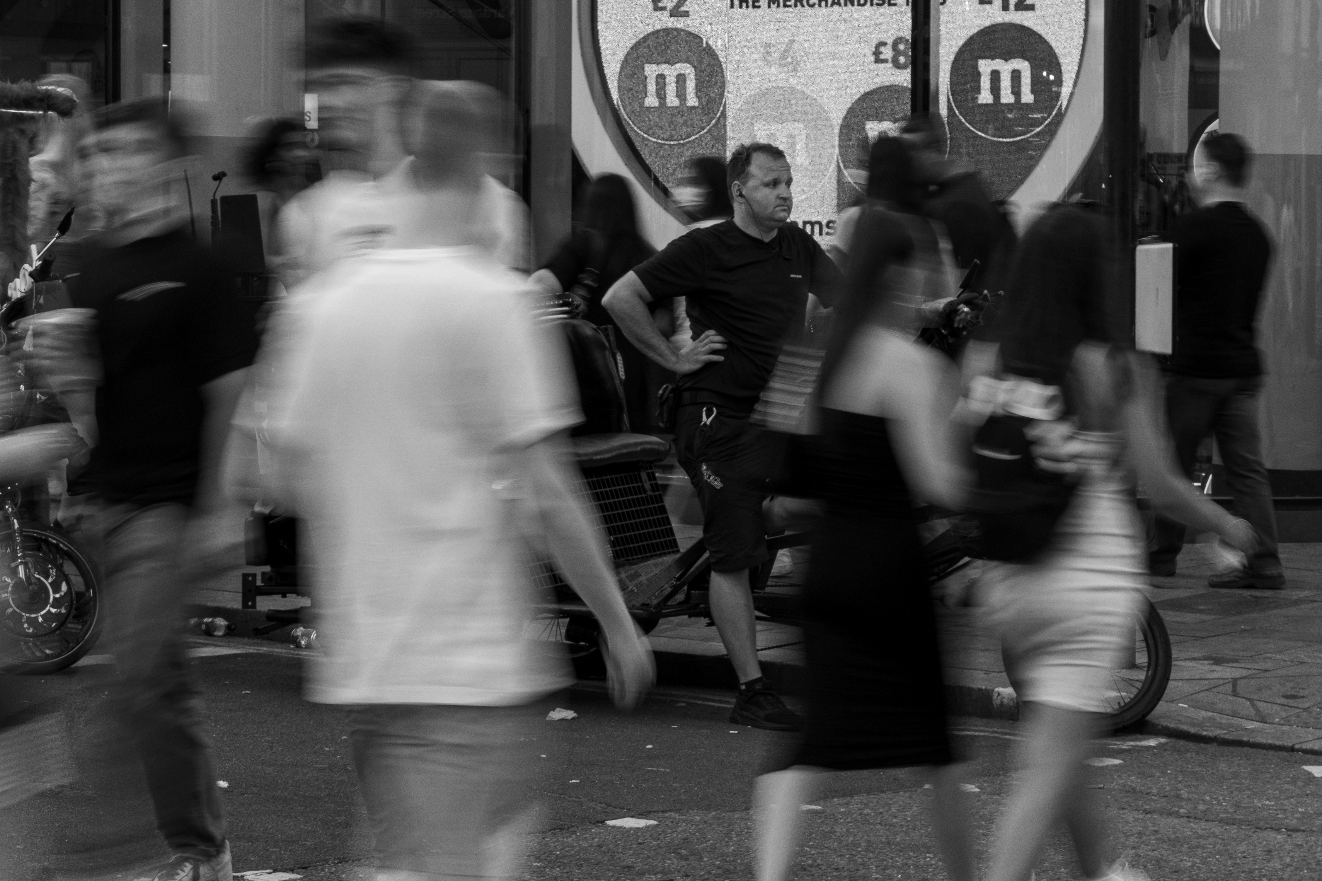 A busy street scene captures the blur of pedestrians moving past a stationary figure, emphasizing the contrast between stillness and motion in urban life.
