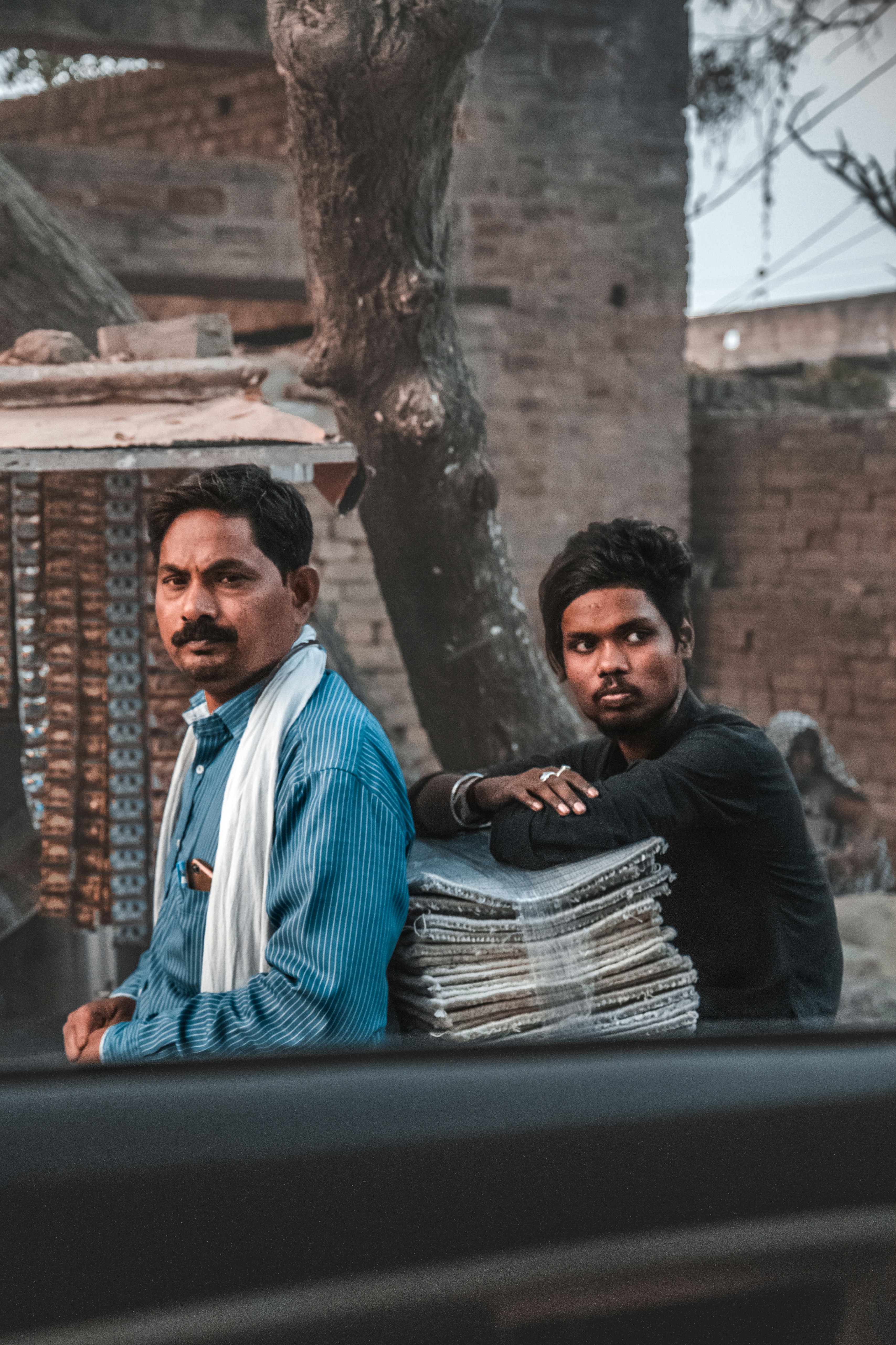 Two men lean on a stack of newspapers beside a tree in a dusty street, captured in muted daylight.