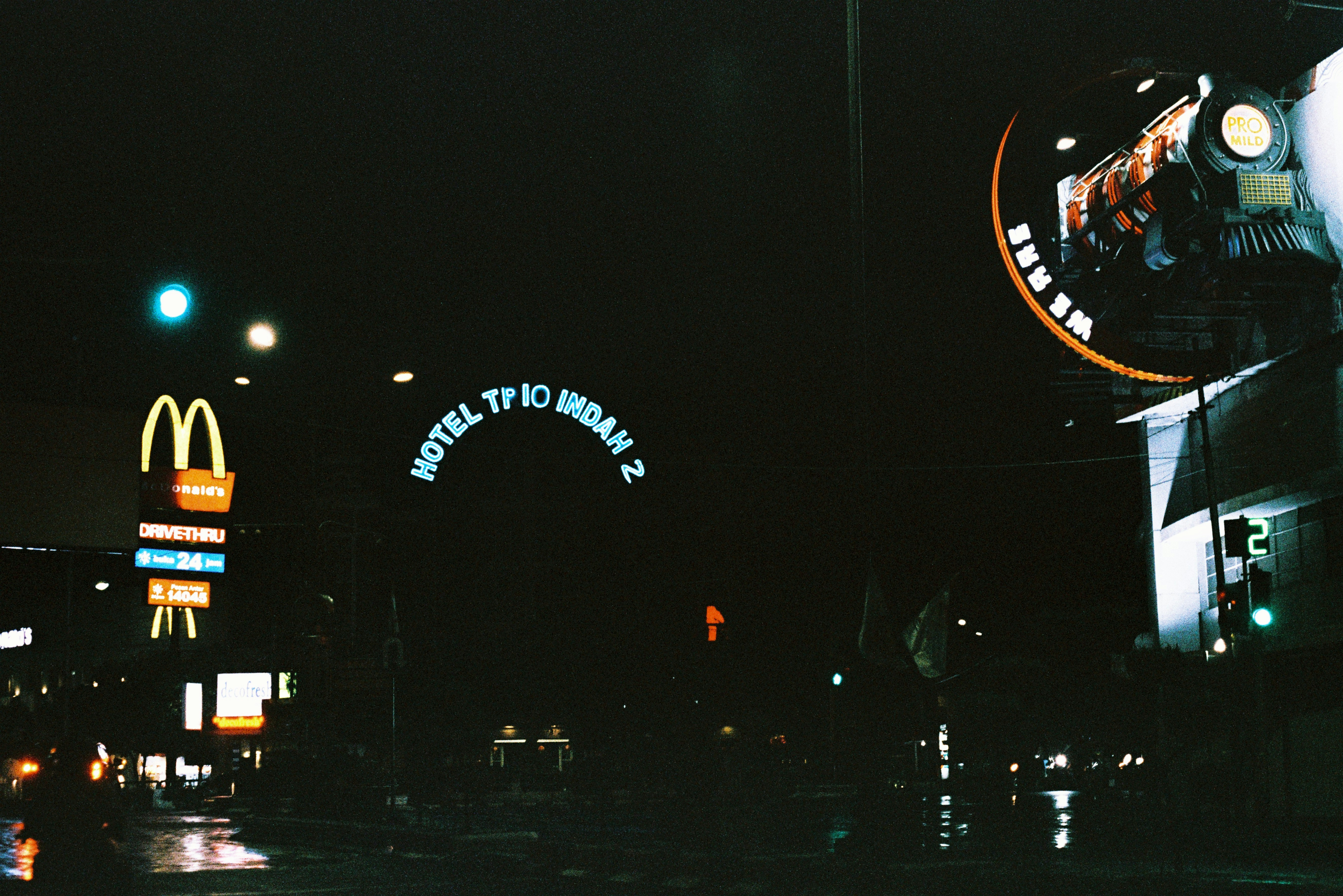 Night-time street scene with wet pavement reflecting neon lights, featuring a McDonald's sign on the left and a curved hotel sign in the center.