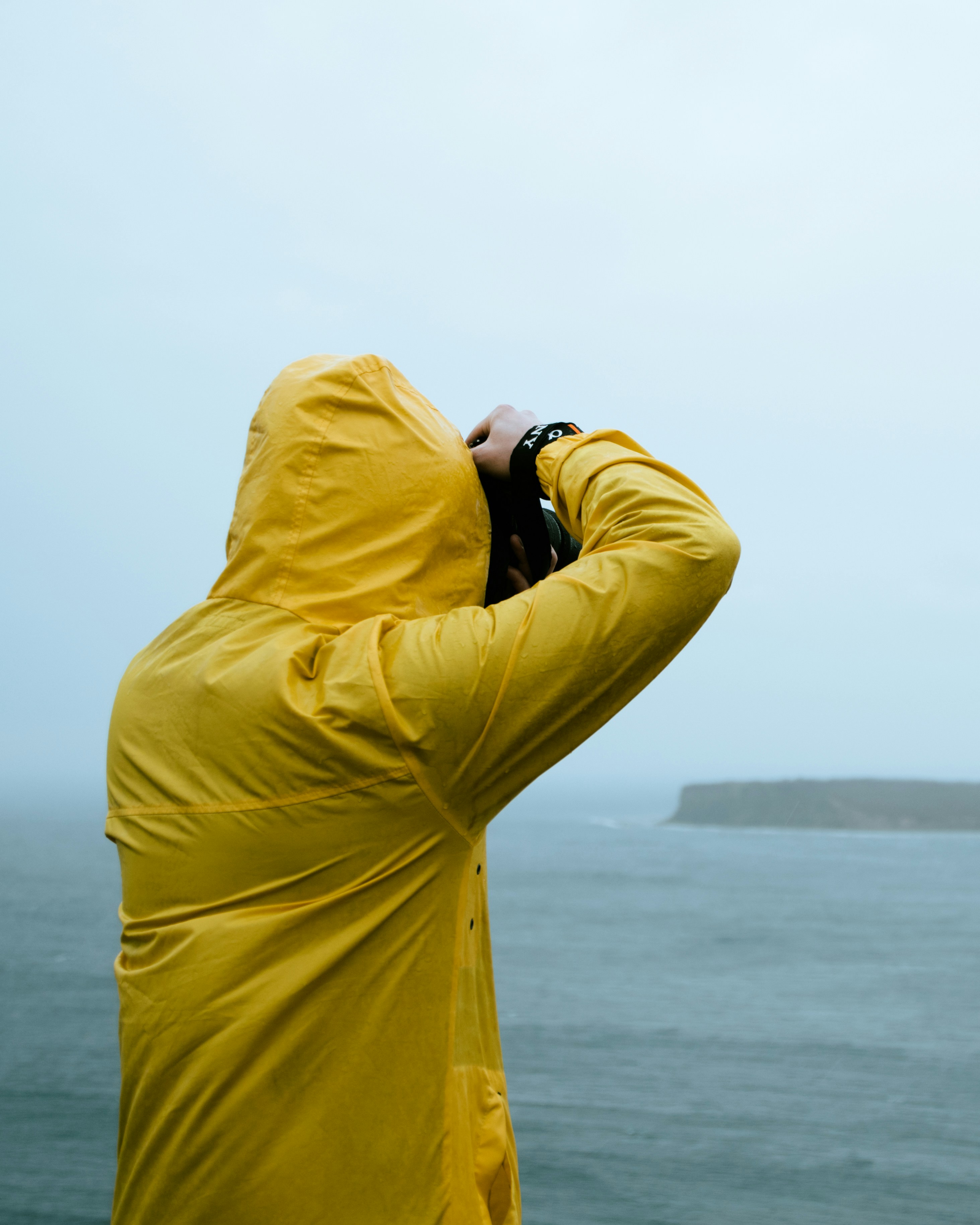 A photographer in a yellow raincoat captures the dramatic coastal landscape amidst a stormy sky. The scene conveys the intensity of nature and the passion for photography.