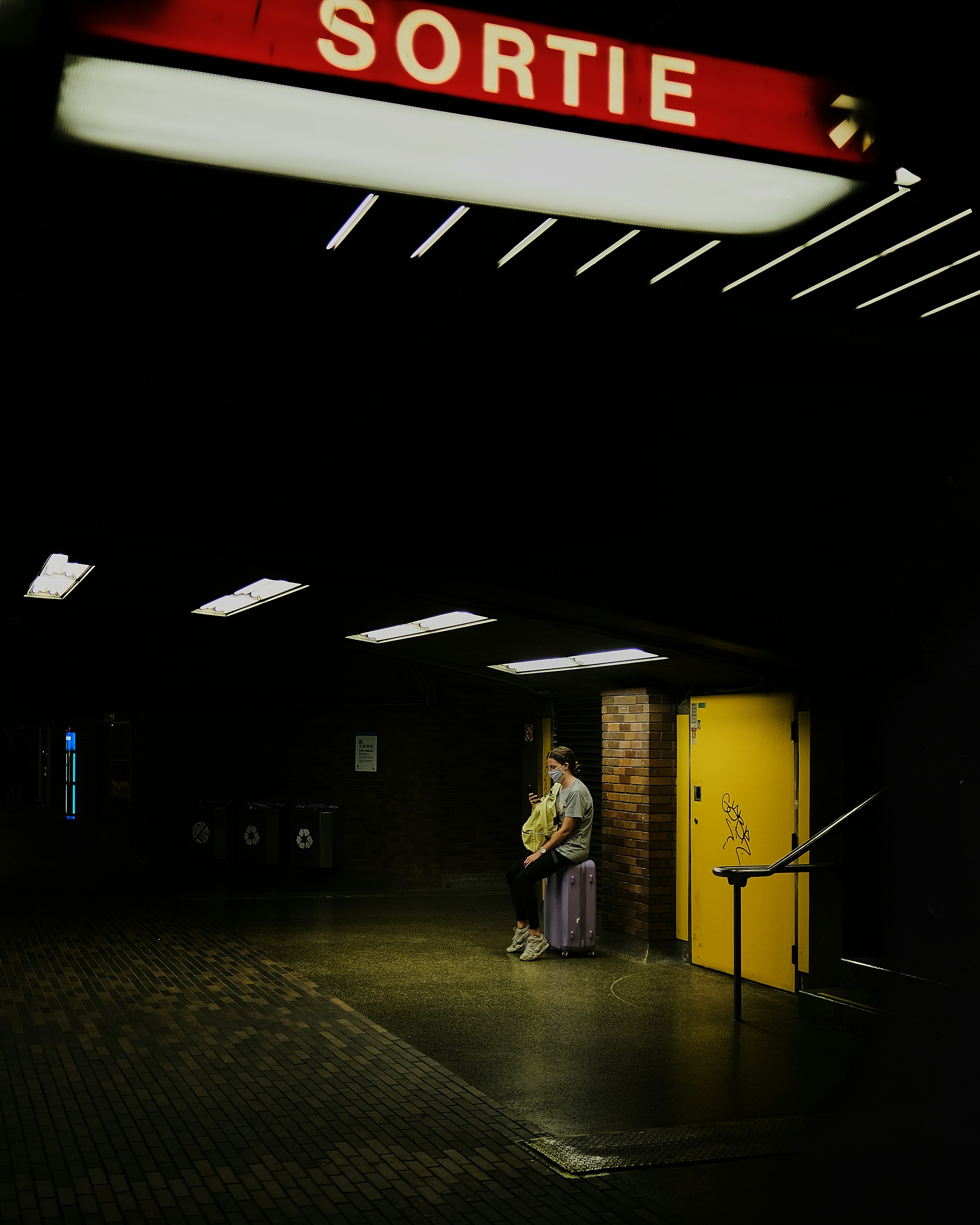 A traveler rests beside a suitcase in a dimly lit subway station, illuminated by overhead lights and a glowing exit sign.