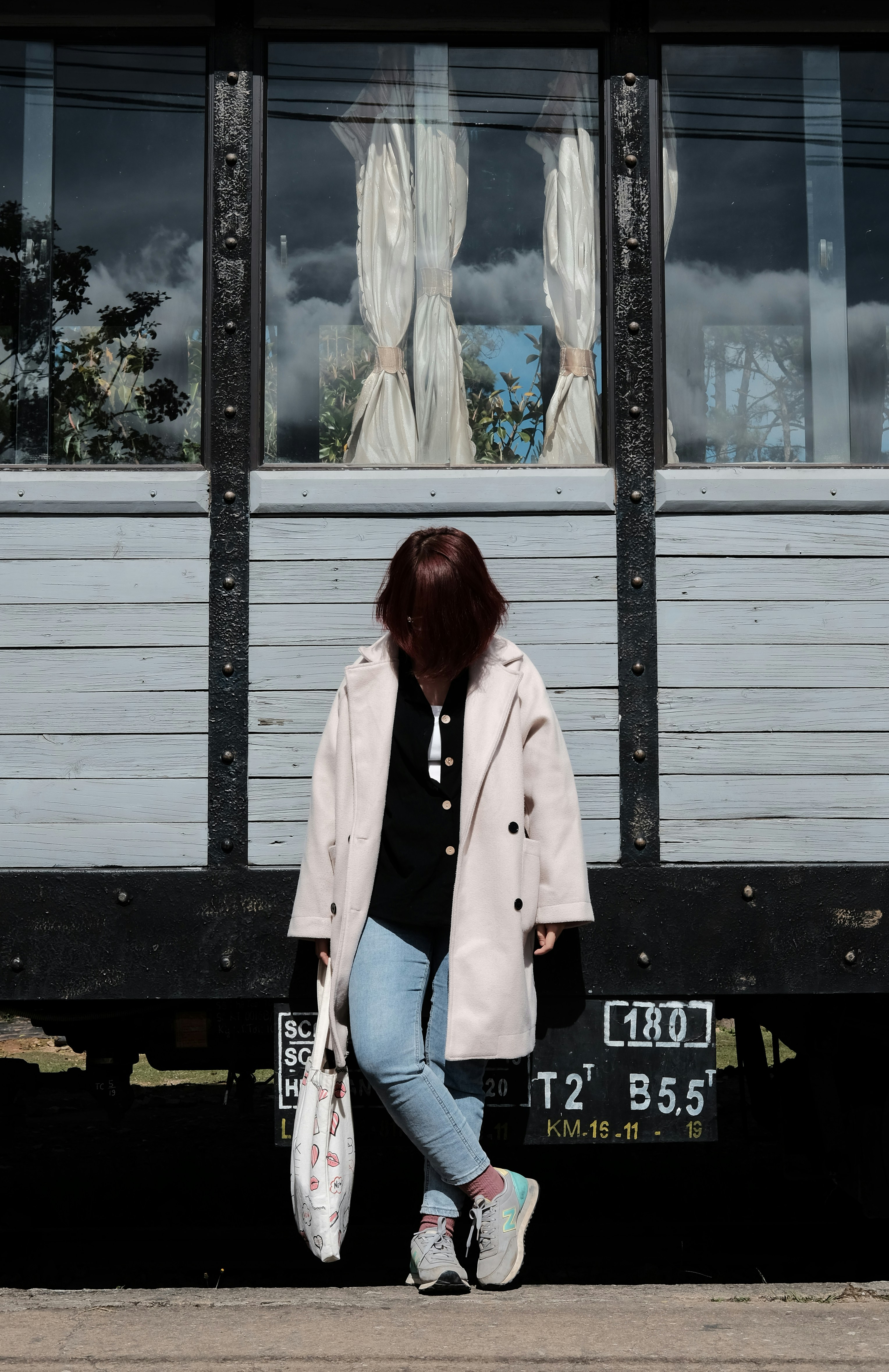 A person stands pensively in front of a vintage train, dressed in a light coat and casual attire, with sunlight illuminating the scene.
