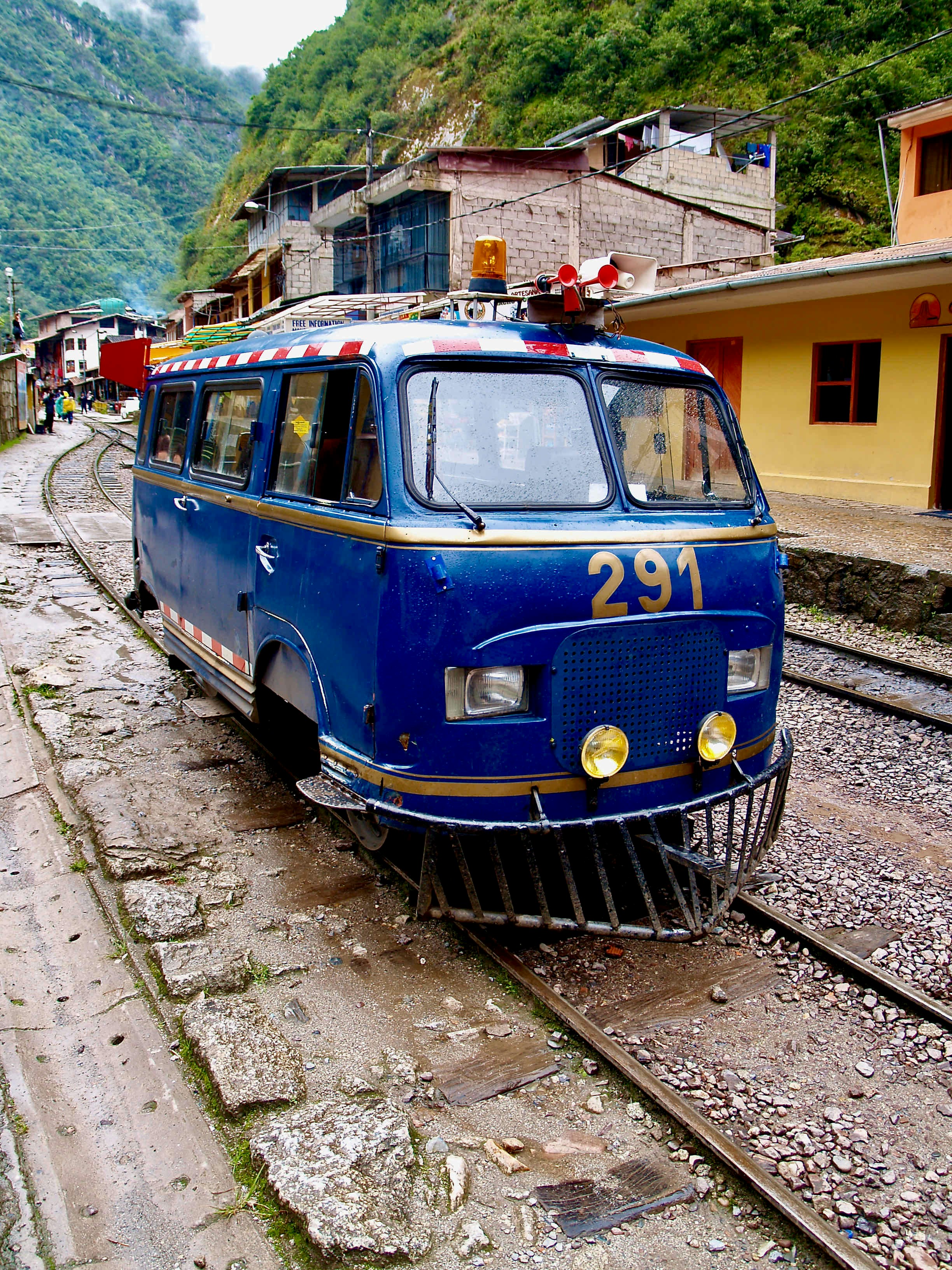 A blue vintage vehicle, identified by the number 291, parked alongside railway tracks in a quaint mountain village, surrounded by lush greenery and rustic buildings.