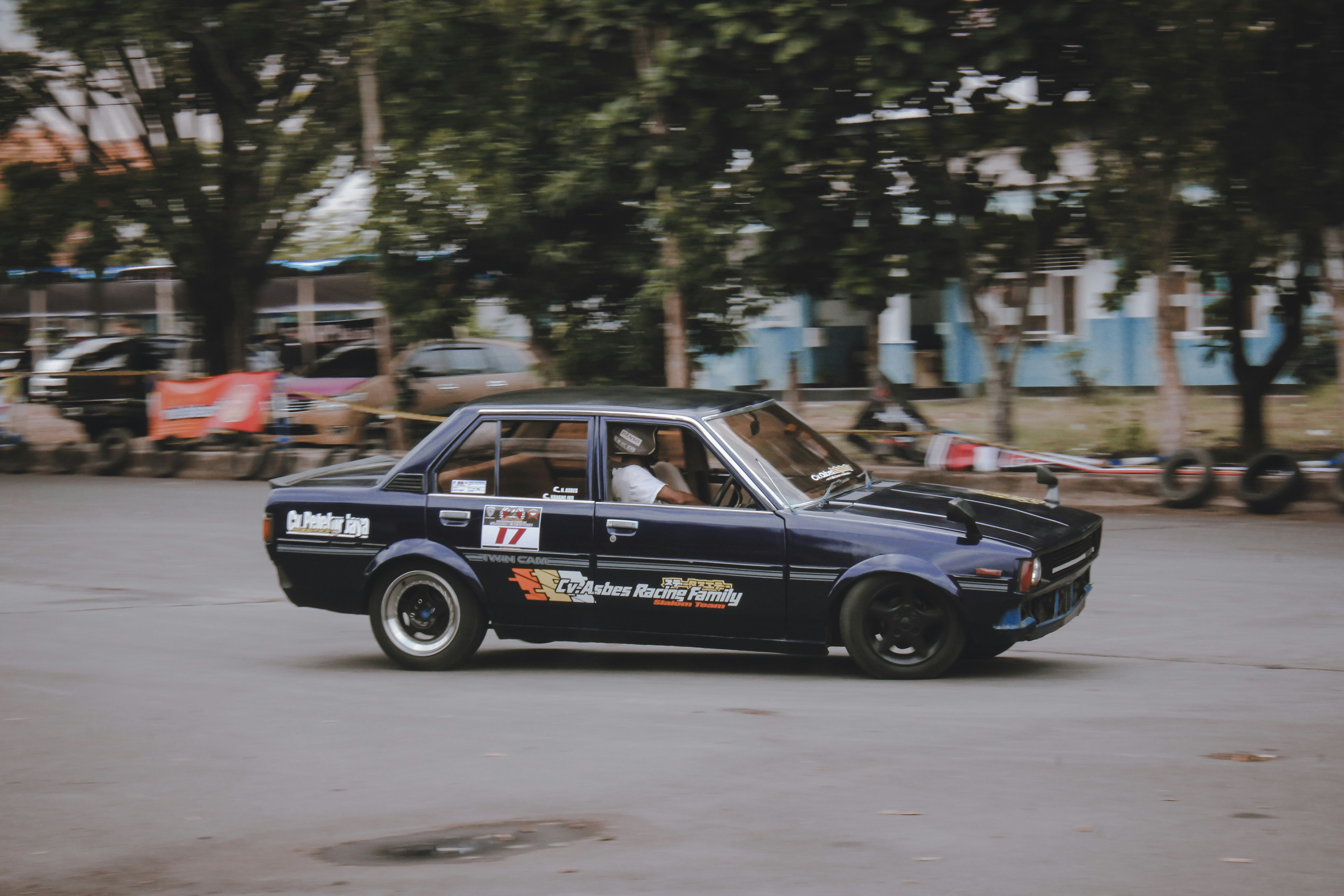 Classic car executing a drift maneuver on an urban street with trees and buildings in the background.